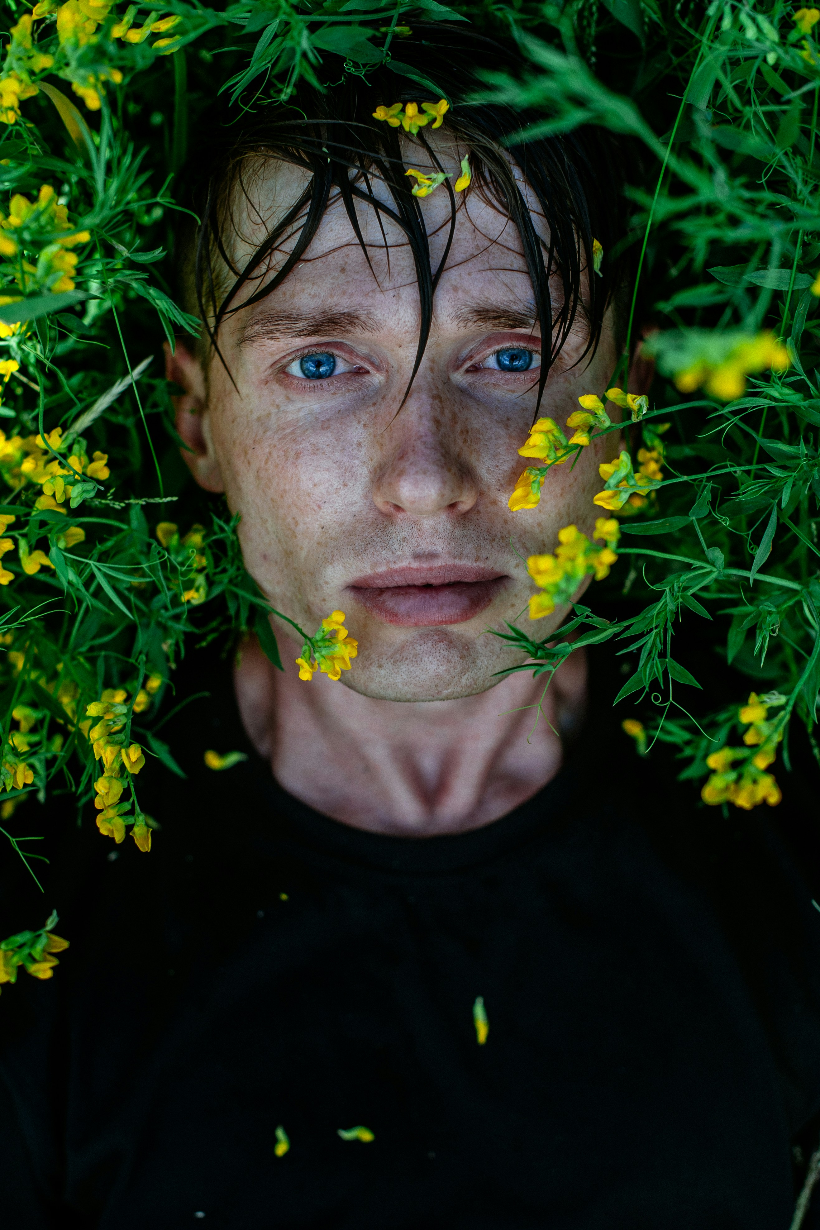 A young man with striking blue eyes surrounded by vibrant yellow flowers and lush green foliage, creating a serene and intimate atmosphere.