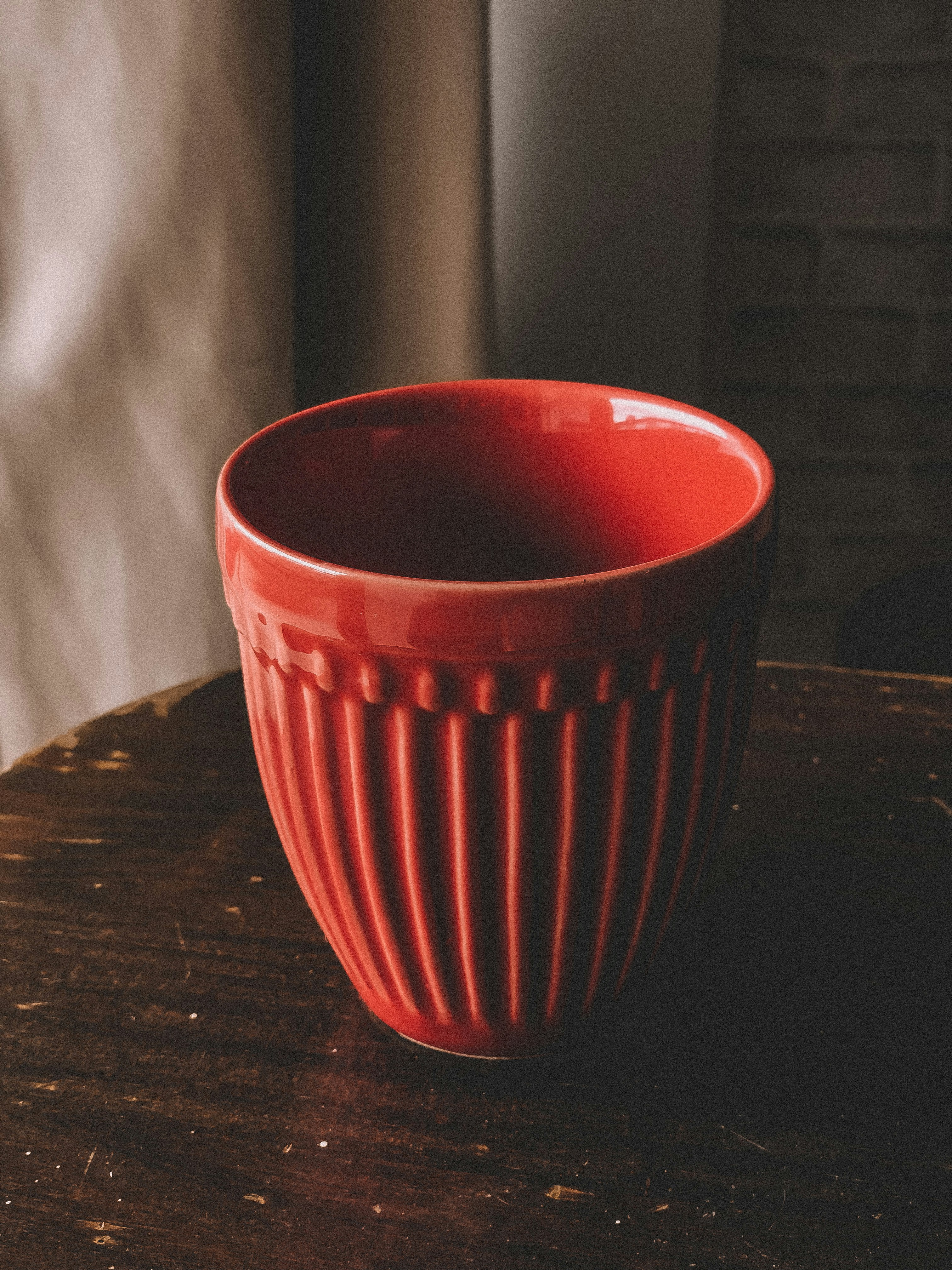 Red ceramic pot with vertical ribbing placed on a wooden table, illuminated by soft natural light.