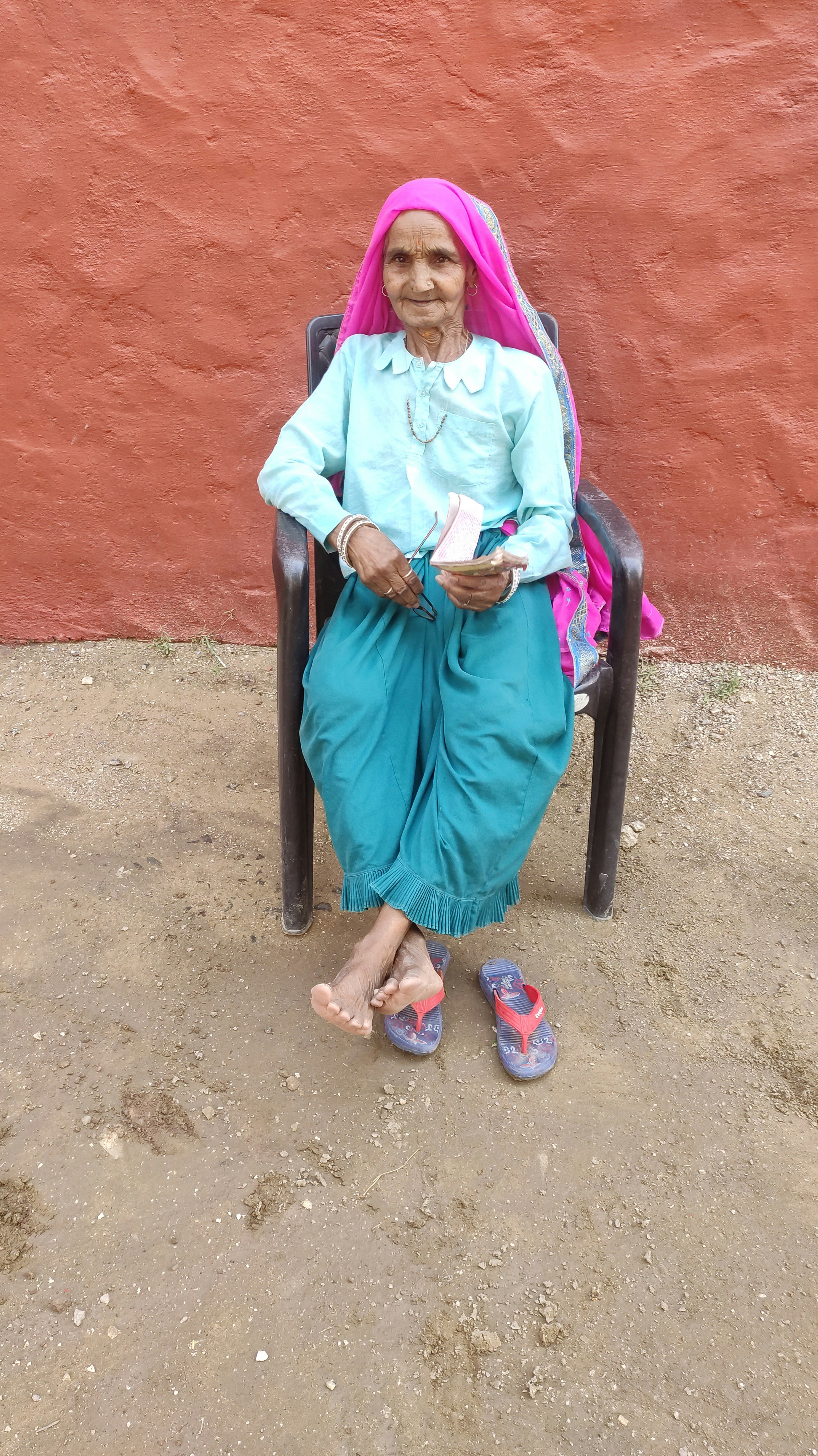 Elderly woman seated on a chair, wearing traditional attire with a colorful scarf, against a rustic red wall. Her expression reflects a lifetime of experiences.