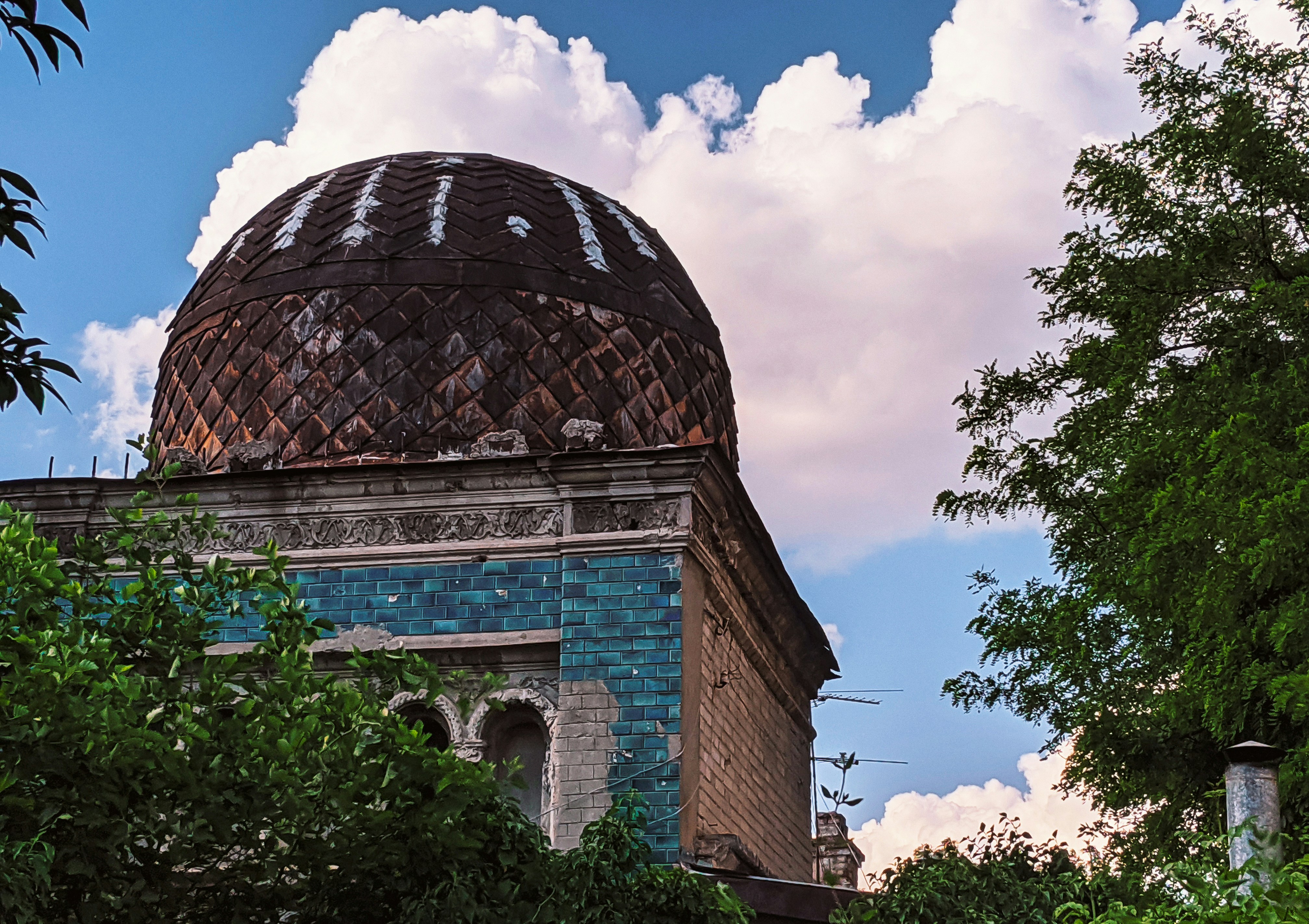 A weathered dome adorned with intricate tiles peeks through lush greenery under a vibrant sky. The remnants of history blend with nature's embrace.