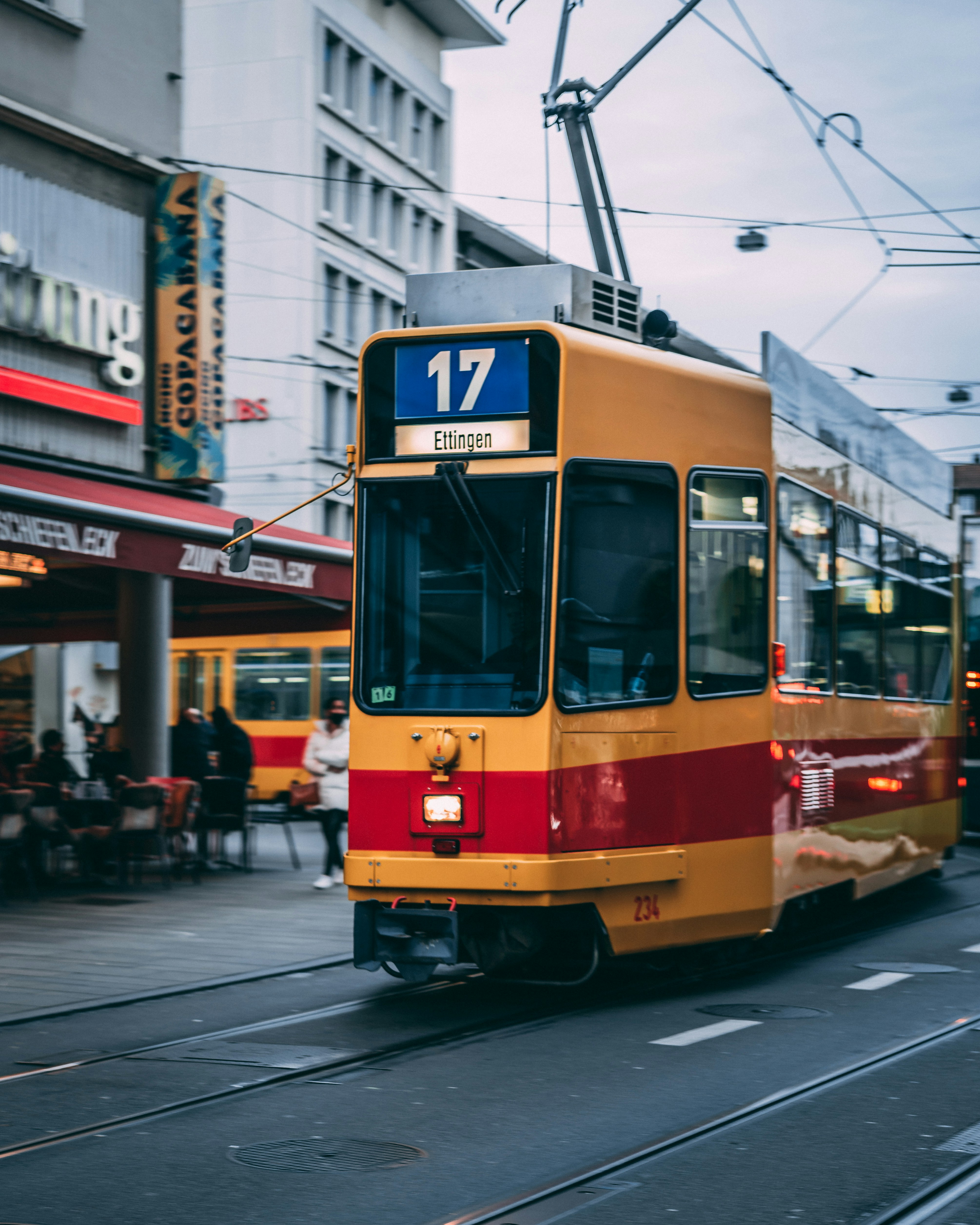 A vibrant yellow tram labeled '17 Ettingen' navigates through a bustling city street, surrounded by cafes and urban architecture.
