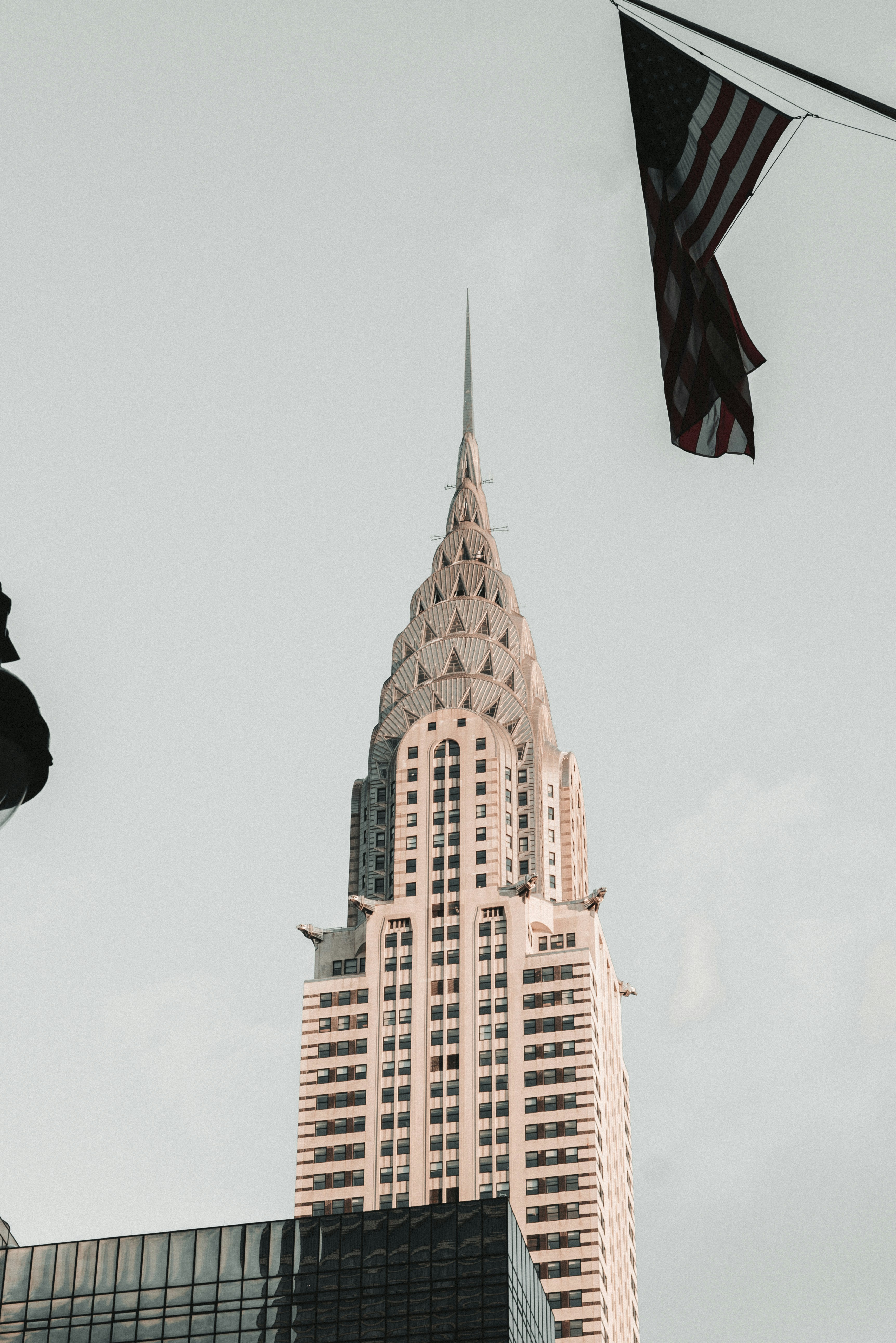 The iconic spire of the Chrysler Building rises majestically against a pale sky, framed by the silhouette of nearby structures and an American flag.