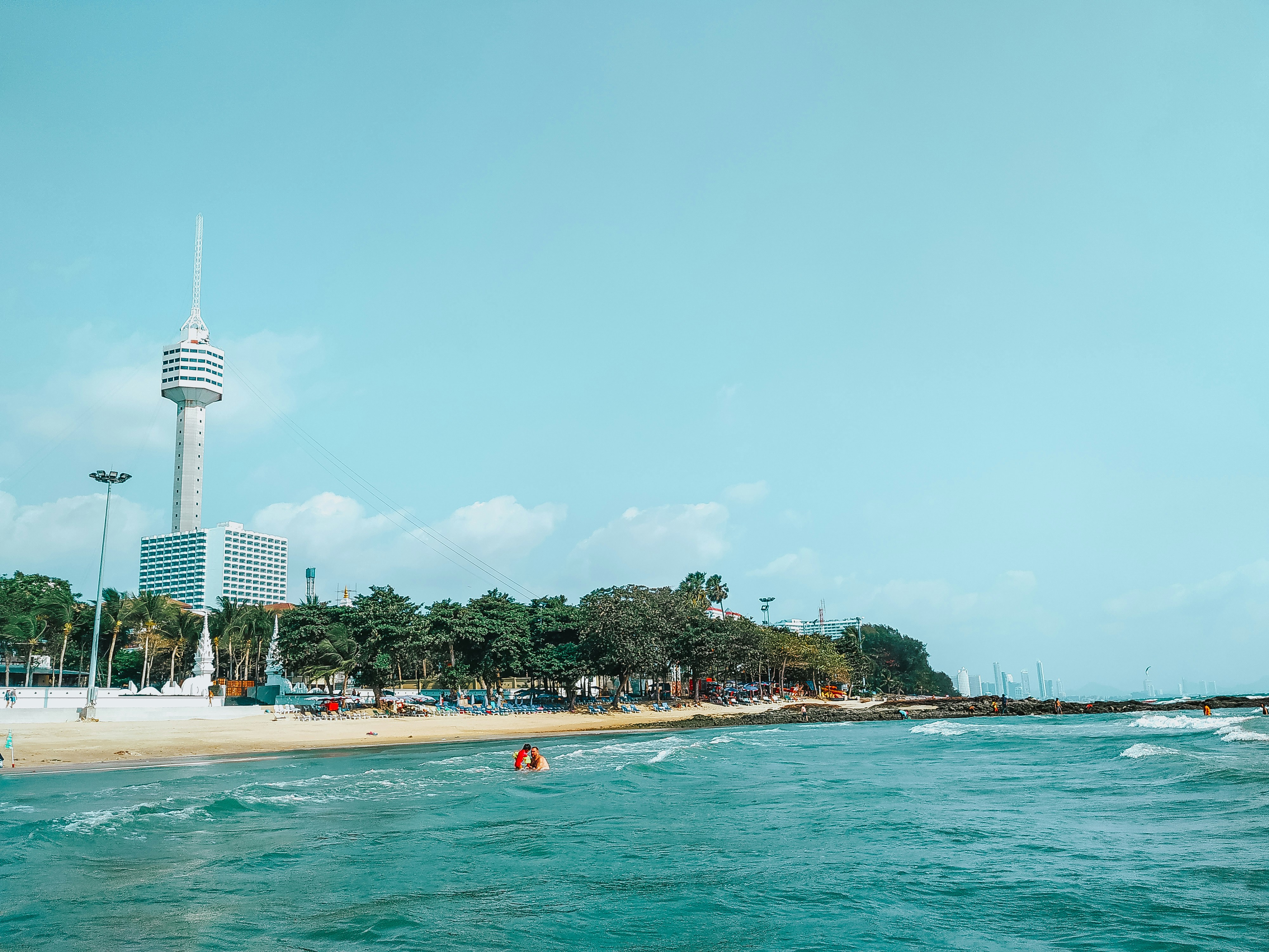 A vibrant beach scene with a prominent tower in the background, showcasing the blend of nature and urban life. The gentle waves lap against the shore under a clear blue sky.
