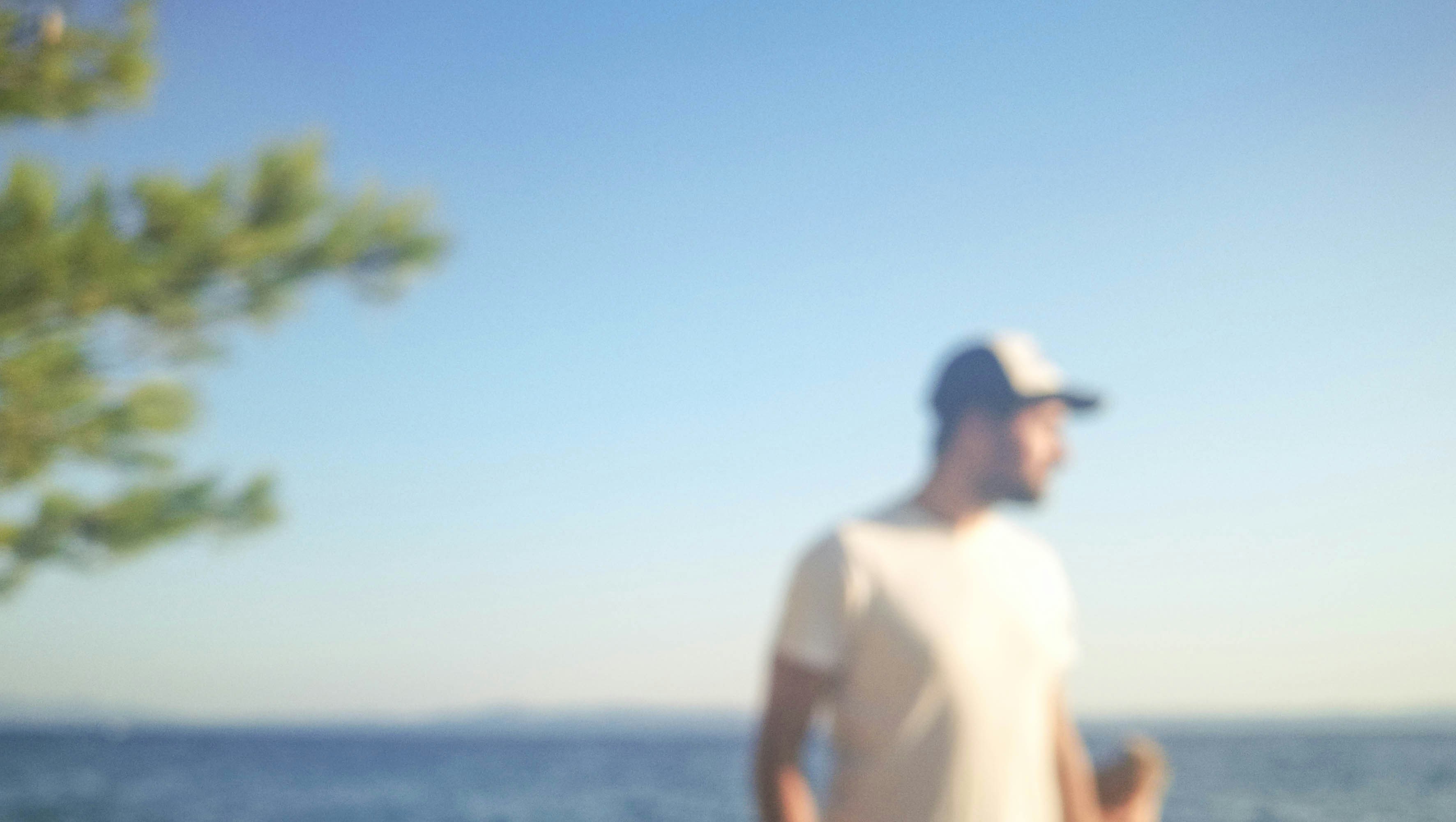 Blurry photograph of a man in a light shirt and cap standing by the sea under a clear blue sky.