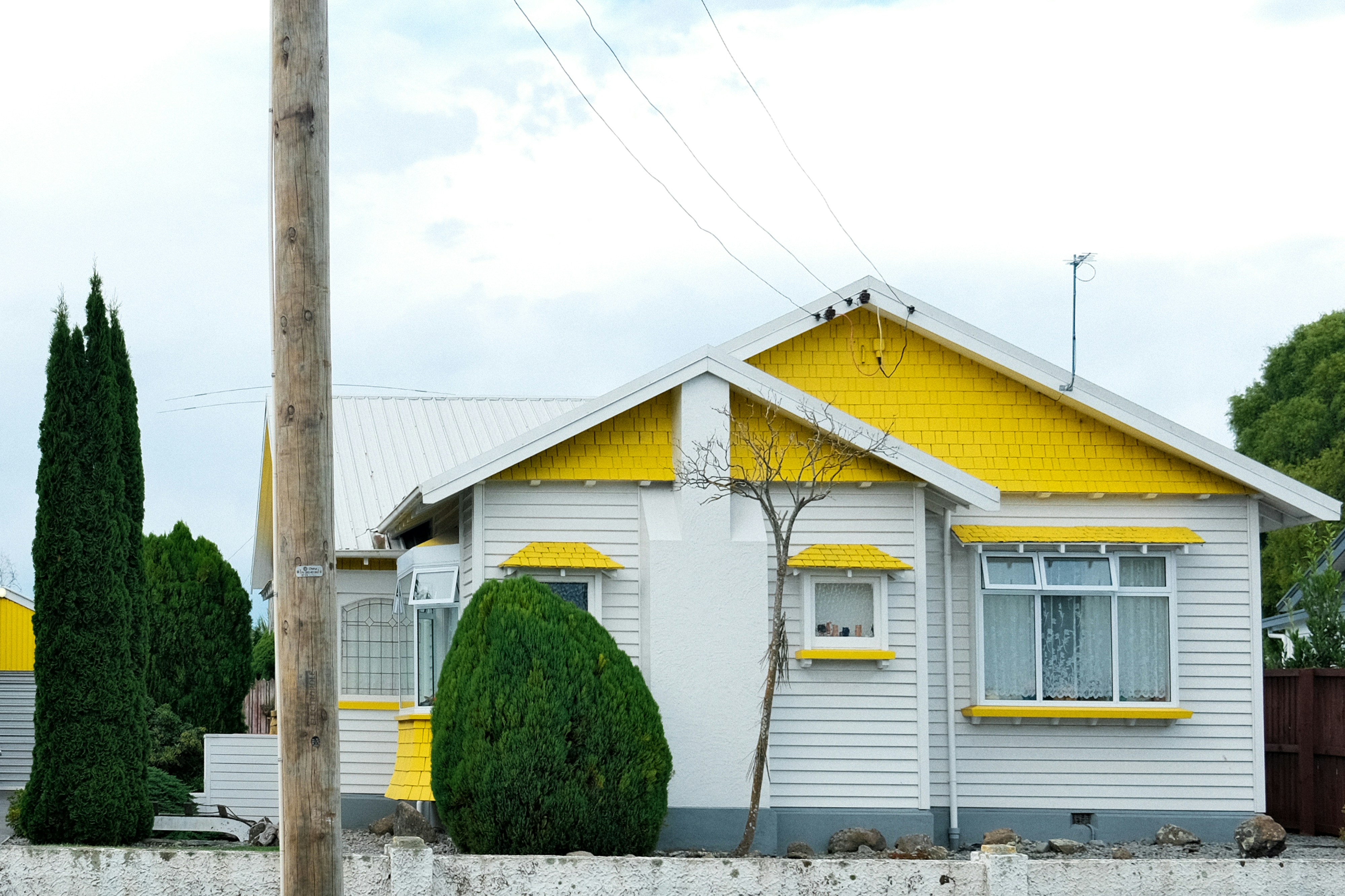 Charming house with vibrant yellow accents, surrounded by lush greenery and power lines, set against a cloudy sky.