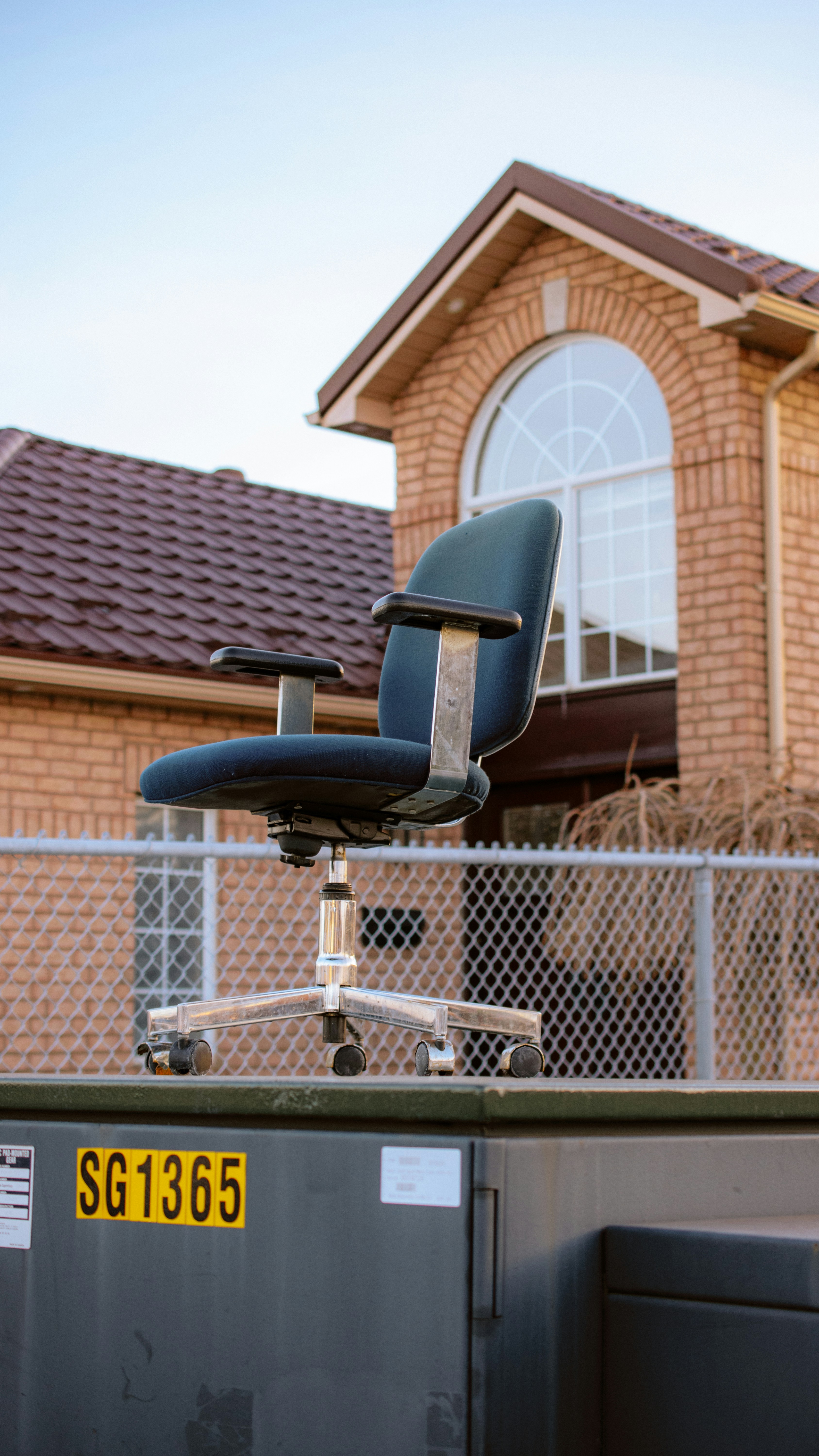 An office chair perched on top of a dumpster, contrasting with a brick house in the background. The scene evokes themes of neglect and unexpected placement.