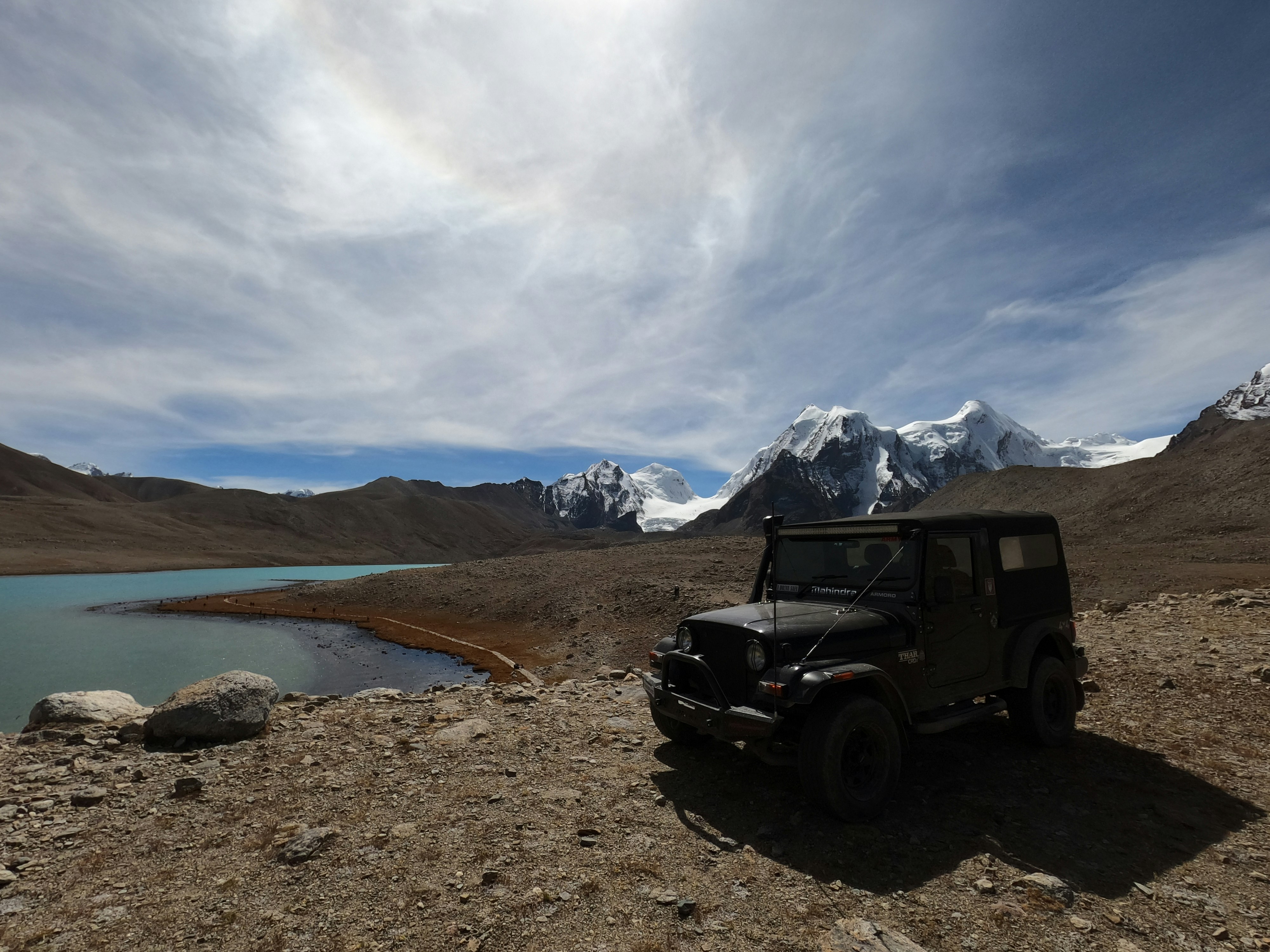 Military-style vehicle parked near a turquoise lake, with snow-capped mountains in the background under a dynamic sky.