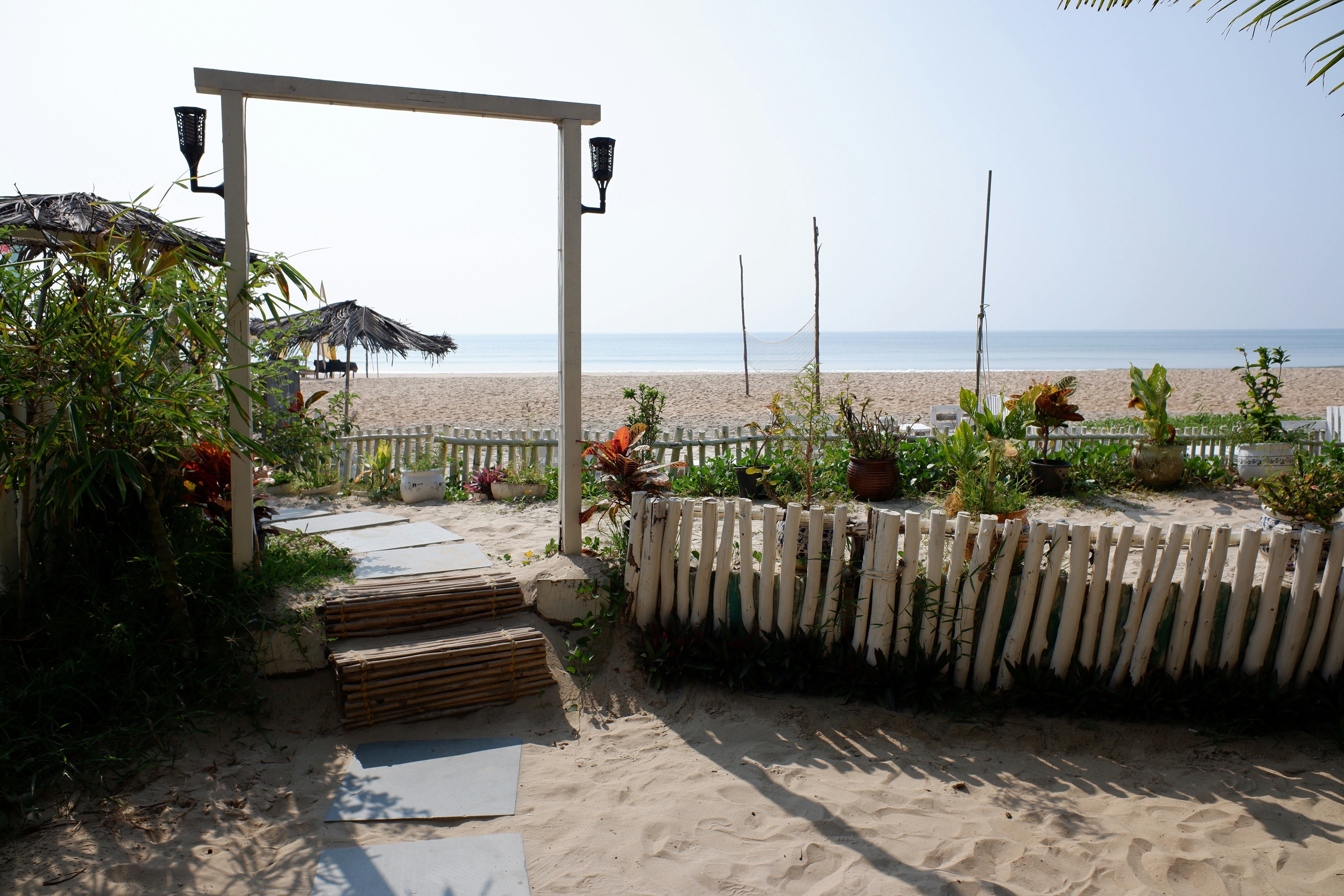 Sunlit beach scene framed by a weathered wooden gate, with steps and a white picket fence leading toward the ocean.
