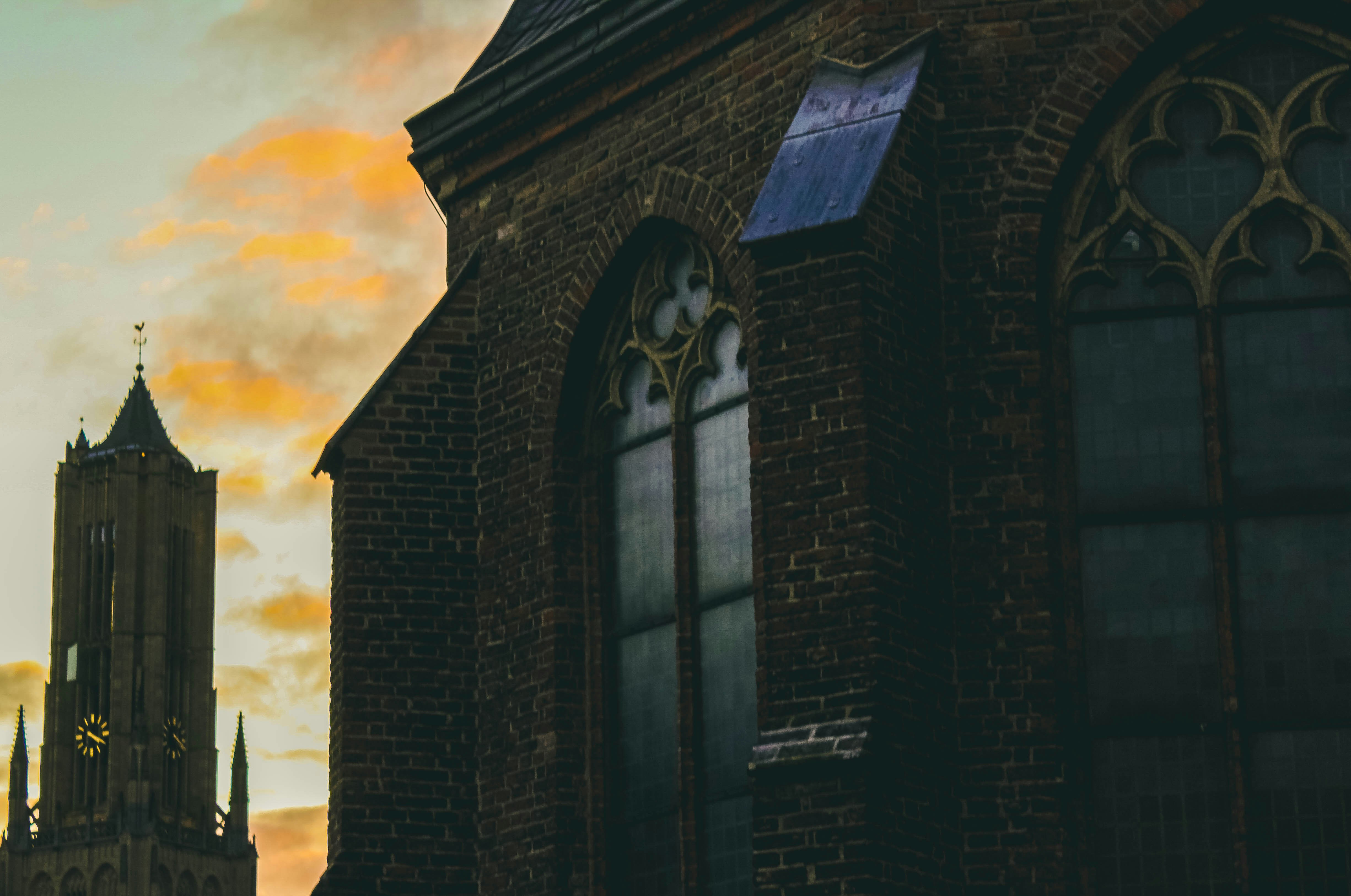Gothic church facade with intricate windows at dusk, with a clock tower in the background against a sunset sky.