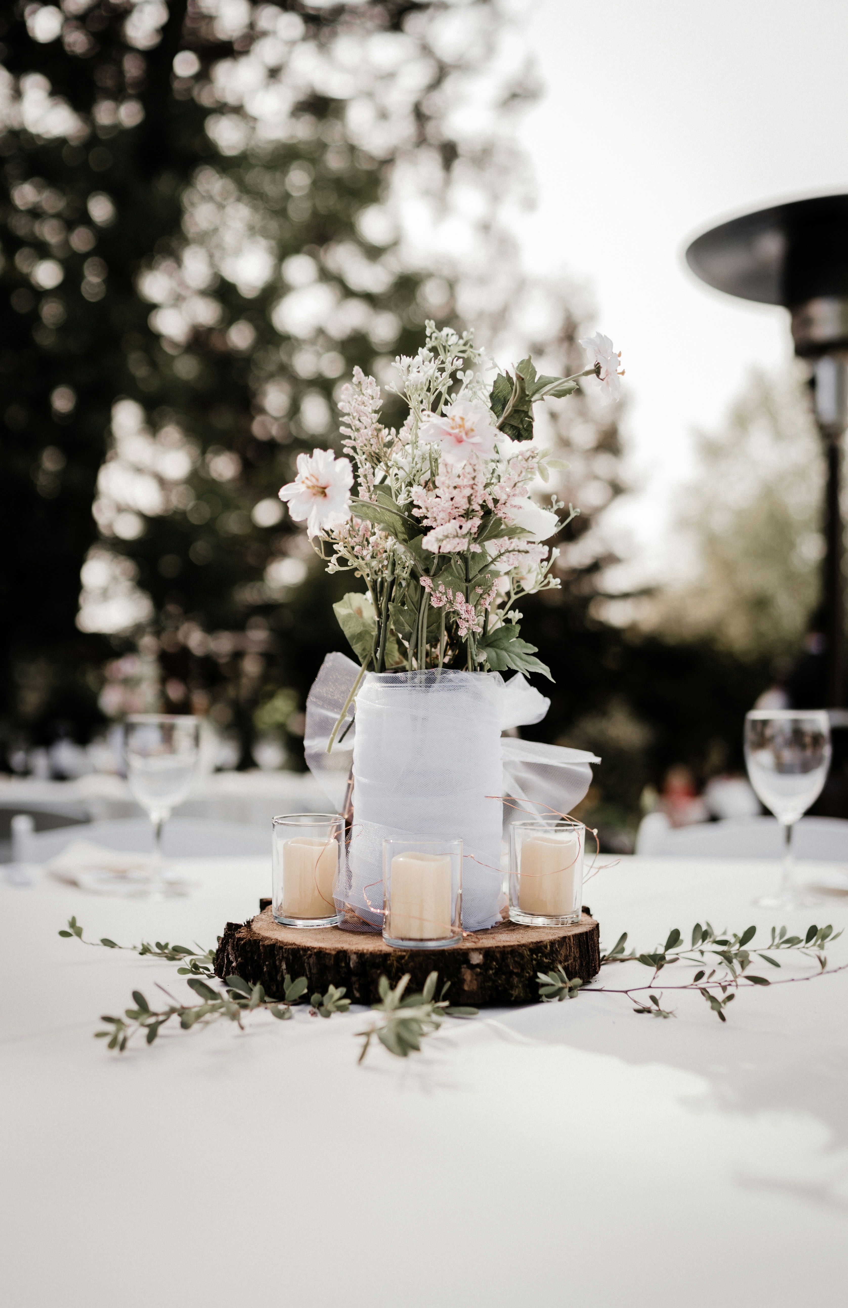 Delicate flowers arranged in a rustic jar atop a wooden slice, surrounded by candles and greenery, creating a charming centerpiece for a festive table setting.