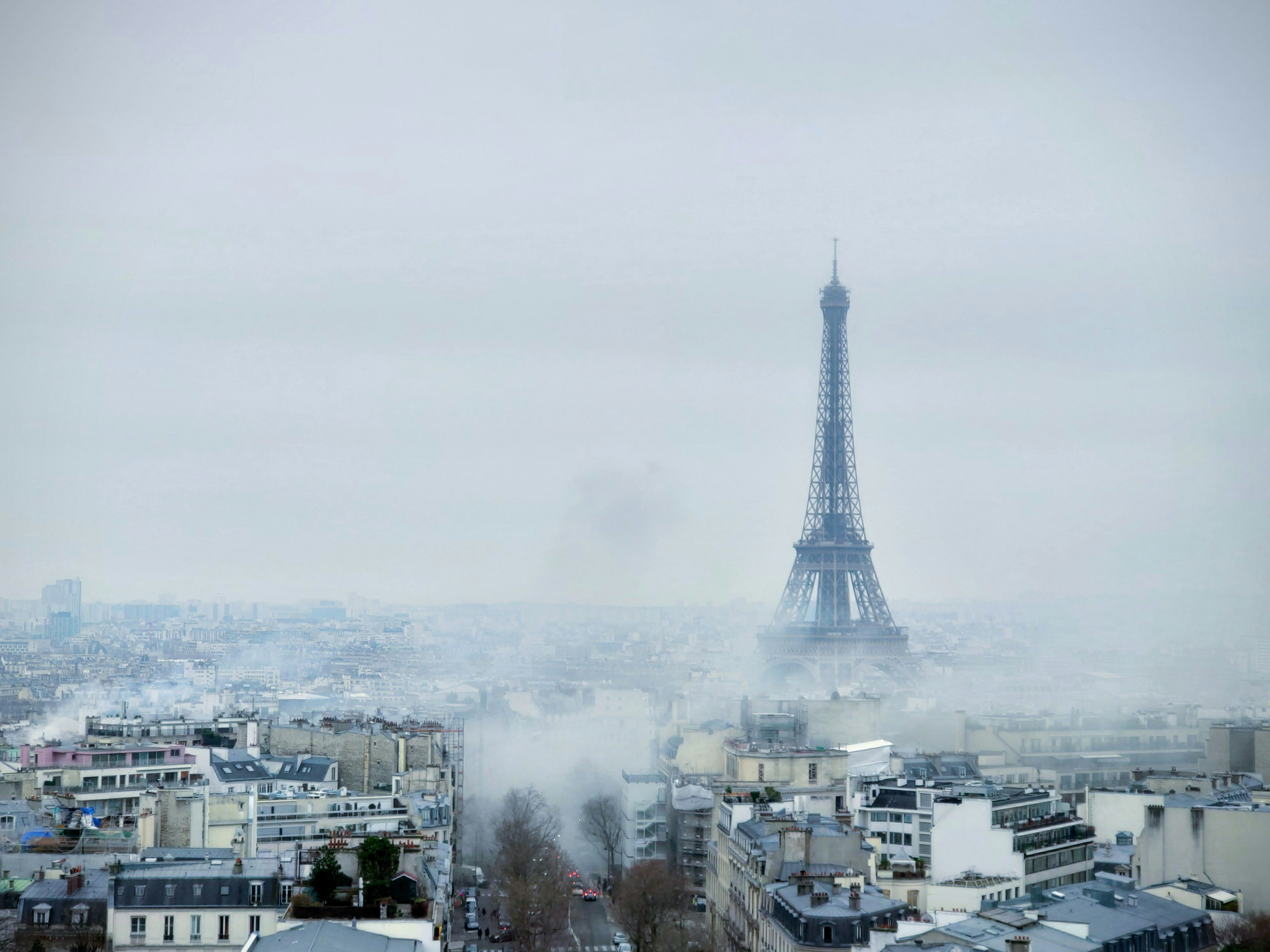 Photograph of the Paris skyline shrouded in pale fog, with the Eiffel Tower rising above the distant rooftops.