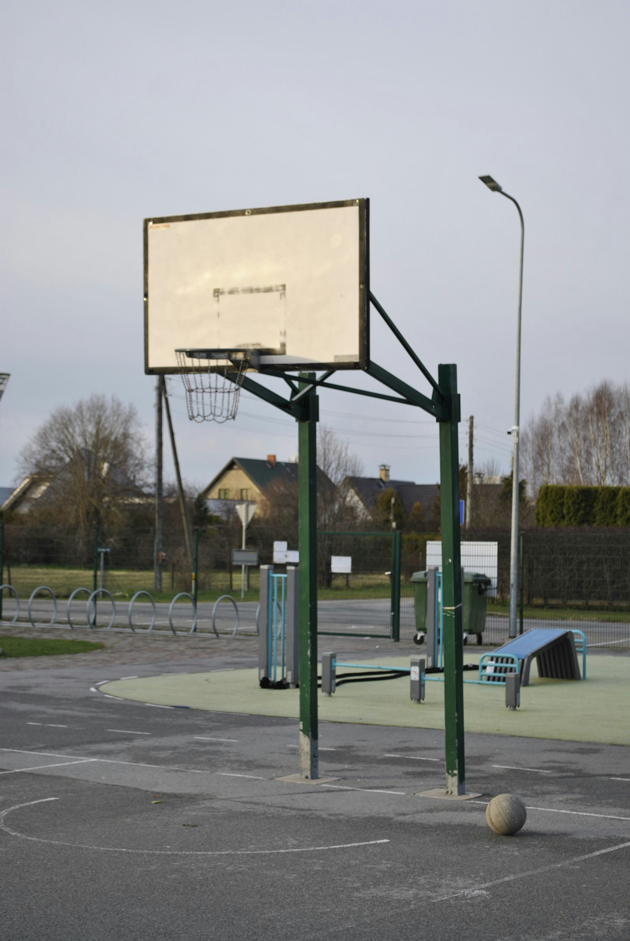 Empty basketball court with a lone ball resting on the asphalt beneath a weathered hoop, hinting at forgotten games and quiet moments of joy.