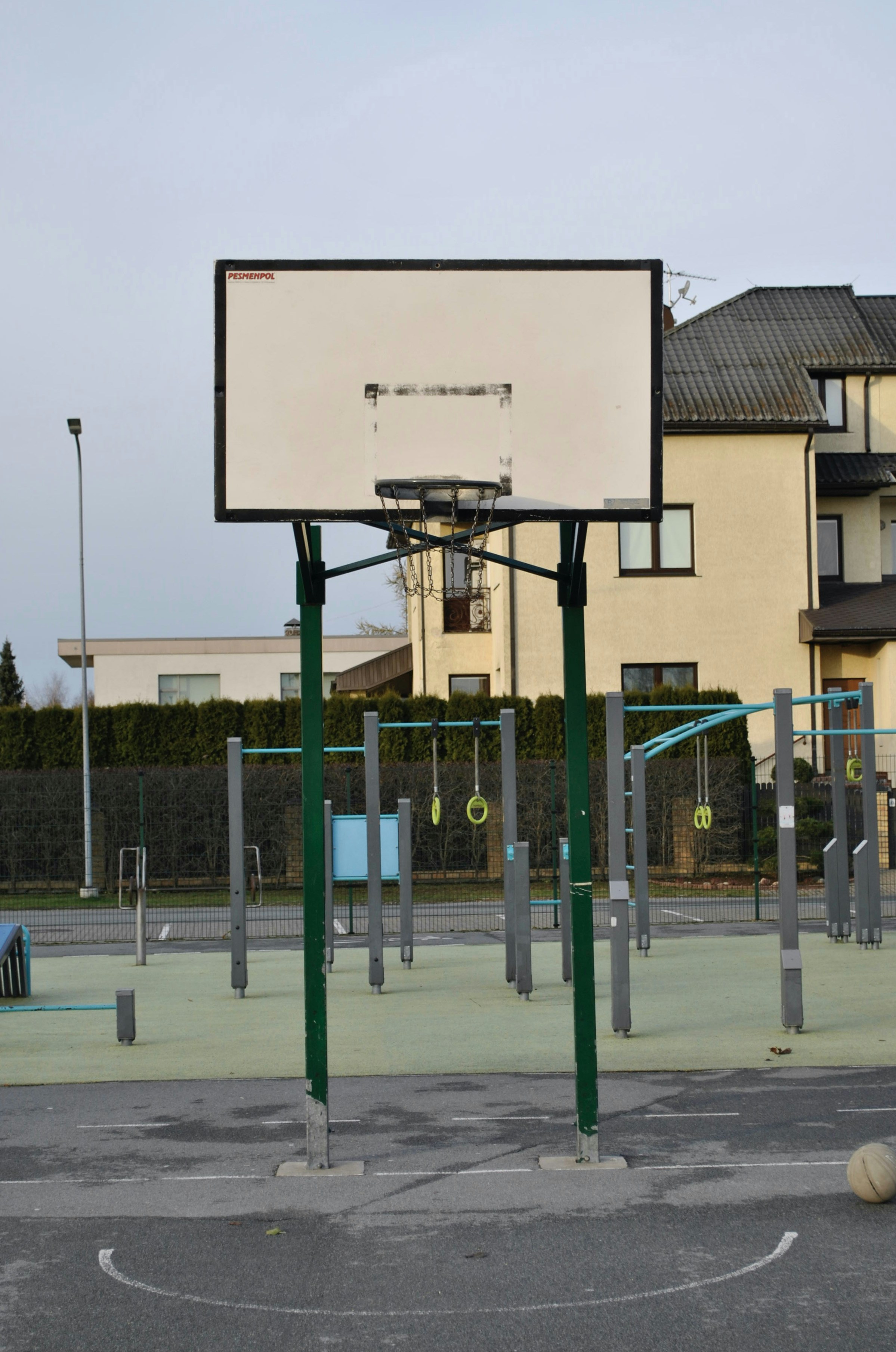 Empty basketball hoop stands prominently in a playground, surrounded by exercise equipment and a quiet neighborhood. 