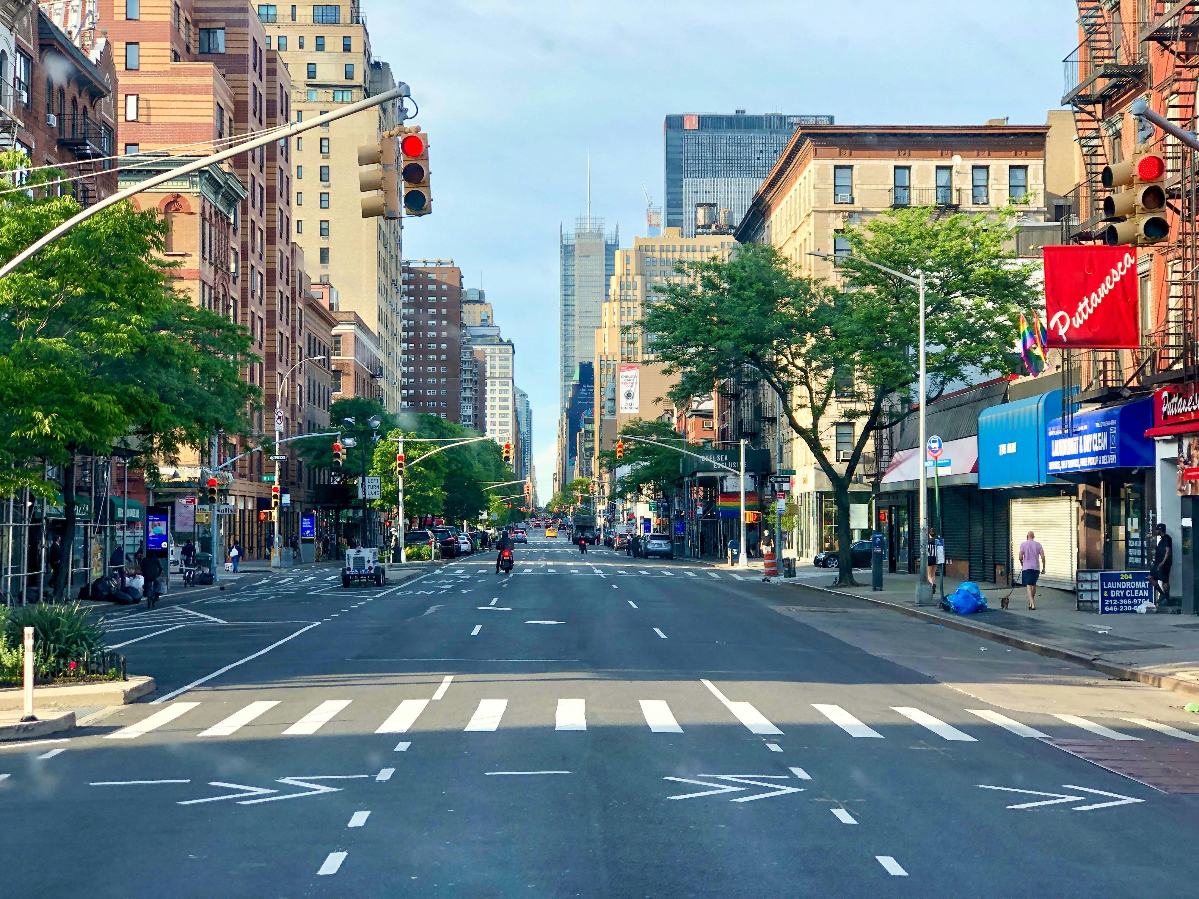 Empty city street flanked by tall buildings and a red traffic light under a clear sky.