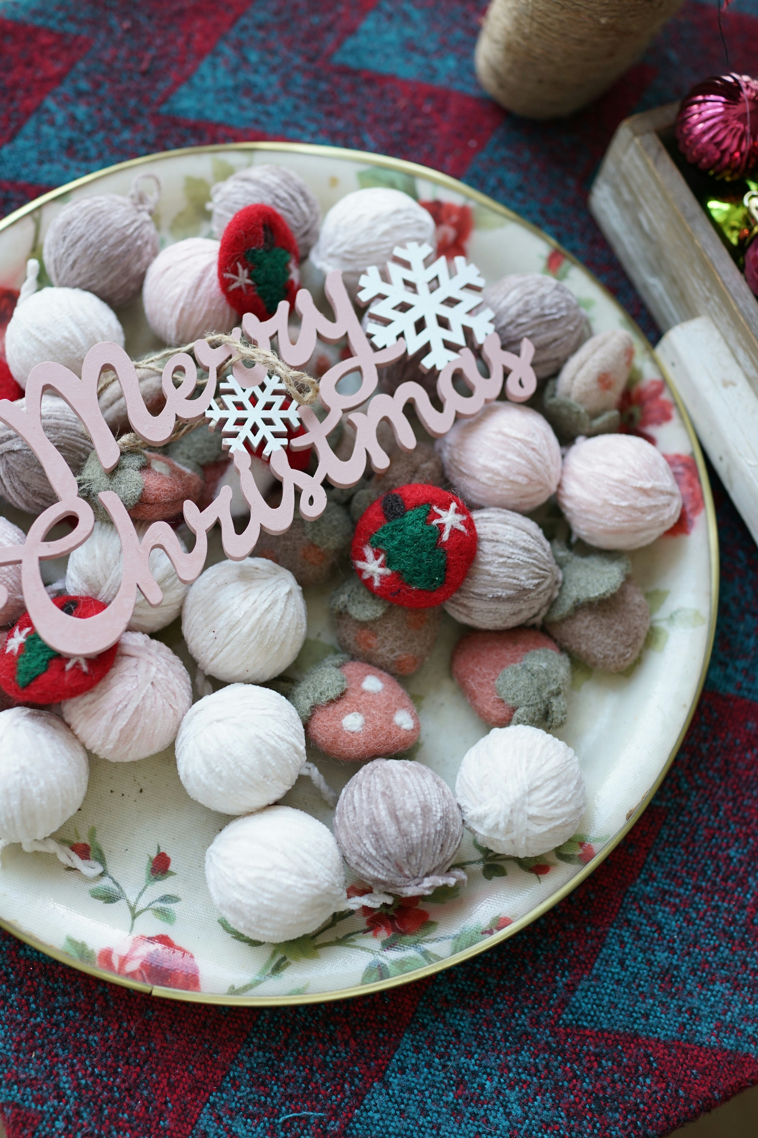Pastel yarn pom-poms fill a floral plate beneath a pink 'Merry Christmas' cutout, creating a festive tabletop arrangement.