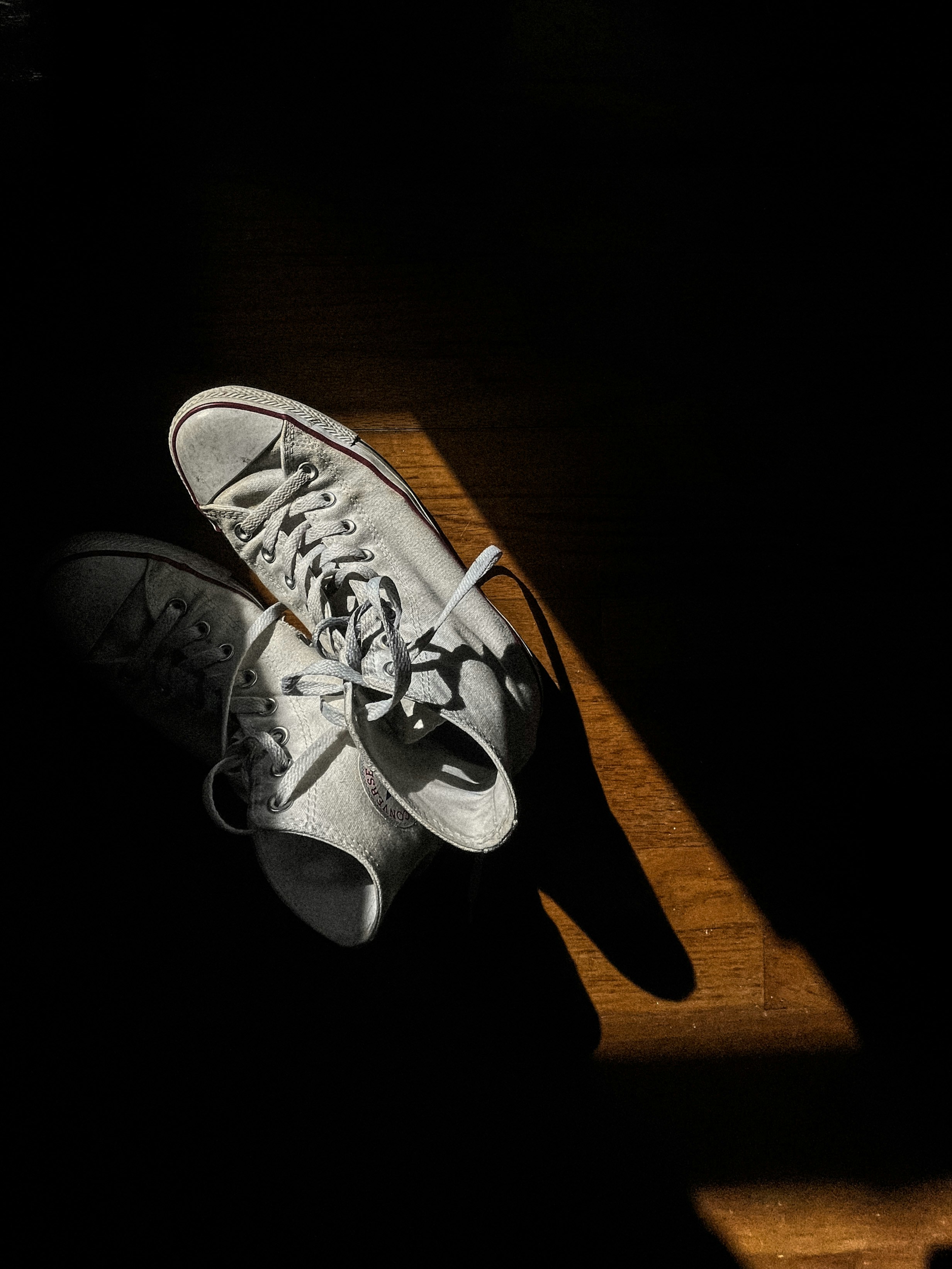 Worn white sneakers resting on a wooden floor, illuminated by a shaft of light casting dramatic shadows.