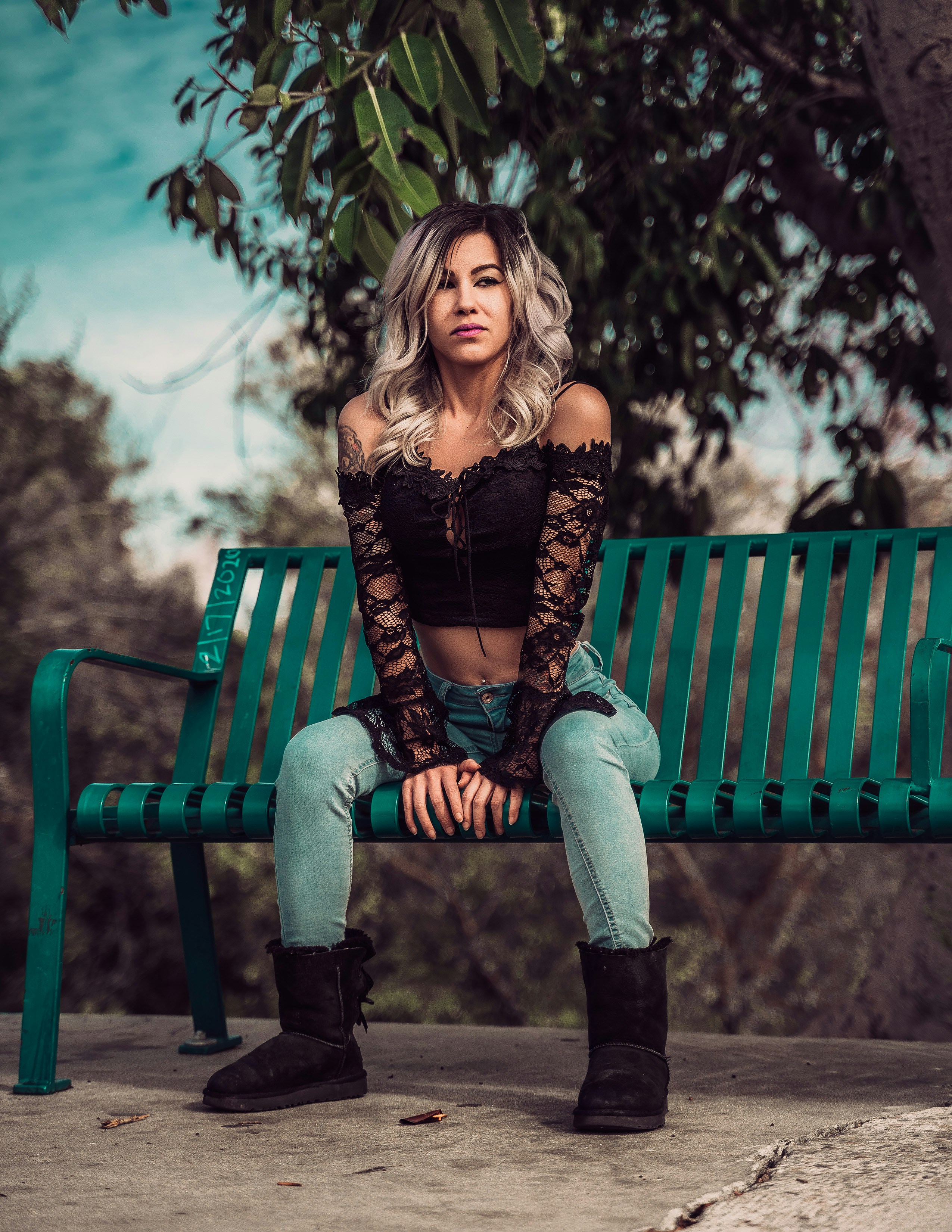 A woman in a black lace top and denim sits thoughtfully on a green bench, surrounded by lush foliage. Her casual yet striking pose conveys a sense of calm amidst urban life.