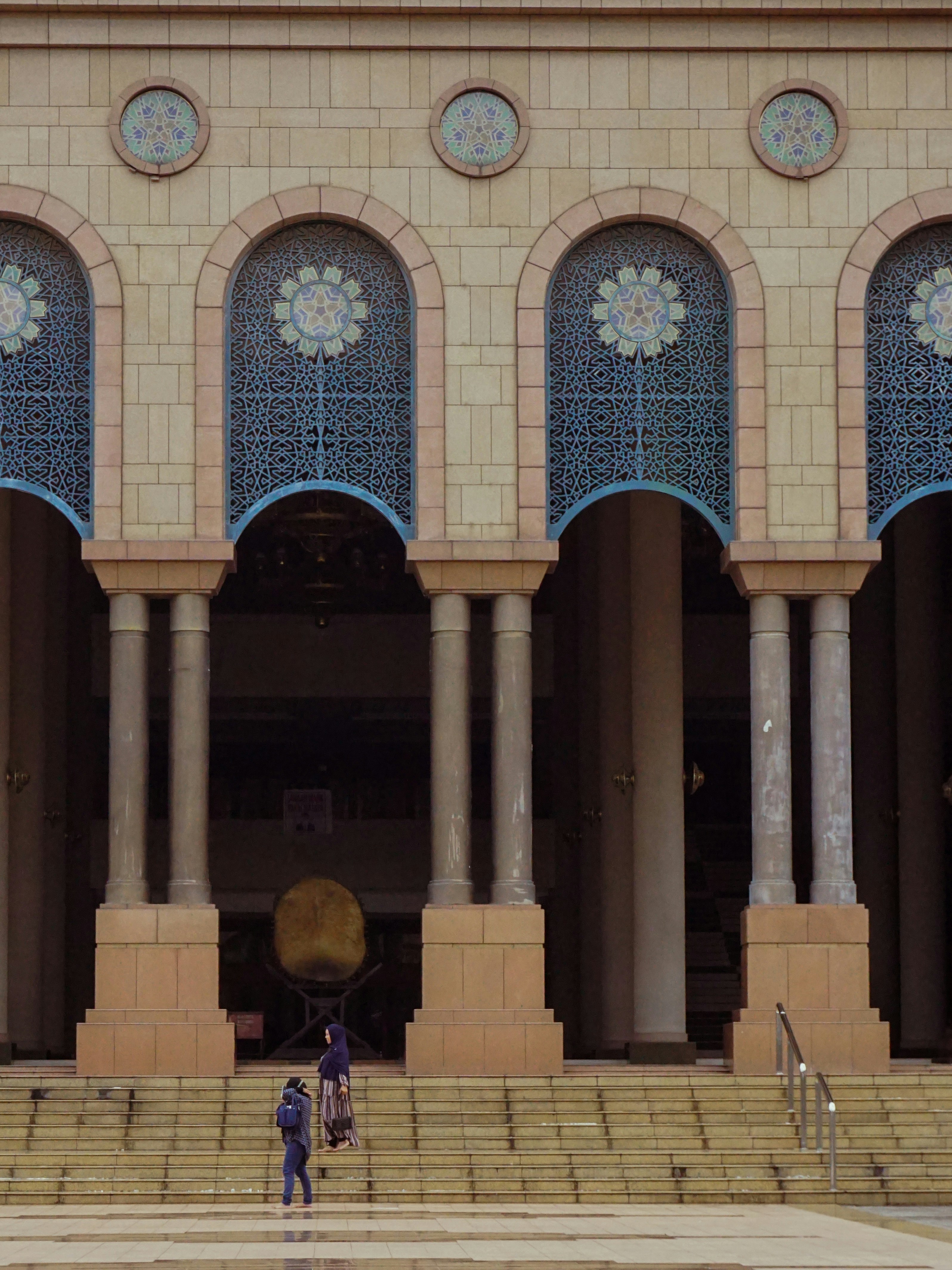 Three people ascend steps toward a grand building with ornate arches and blue decorative panels.