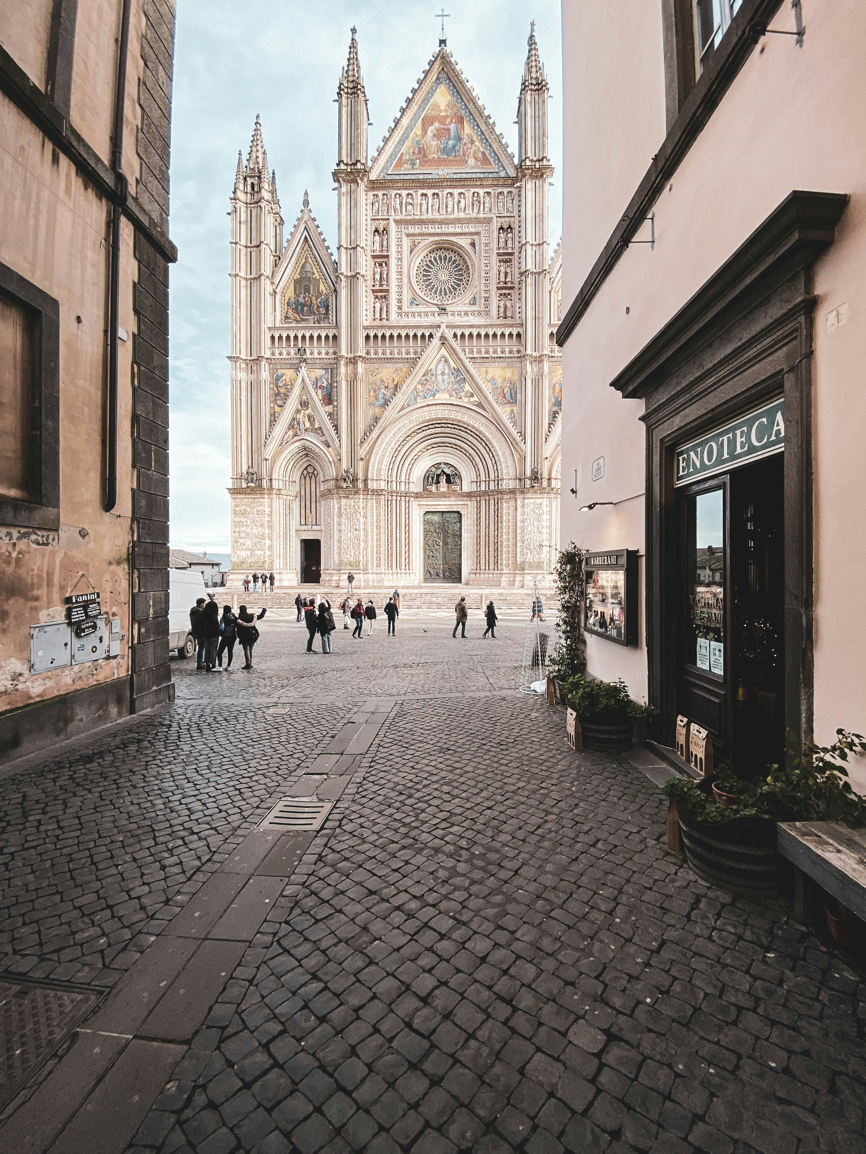 Ornate cathedral rises majestically at the end of a cobblestone street, framed by quaint storefronts. People stroll leisurely, enjoying the historic atmosphere.