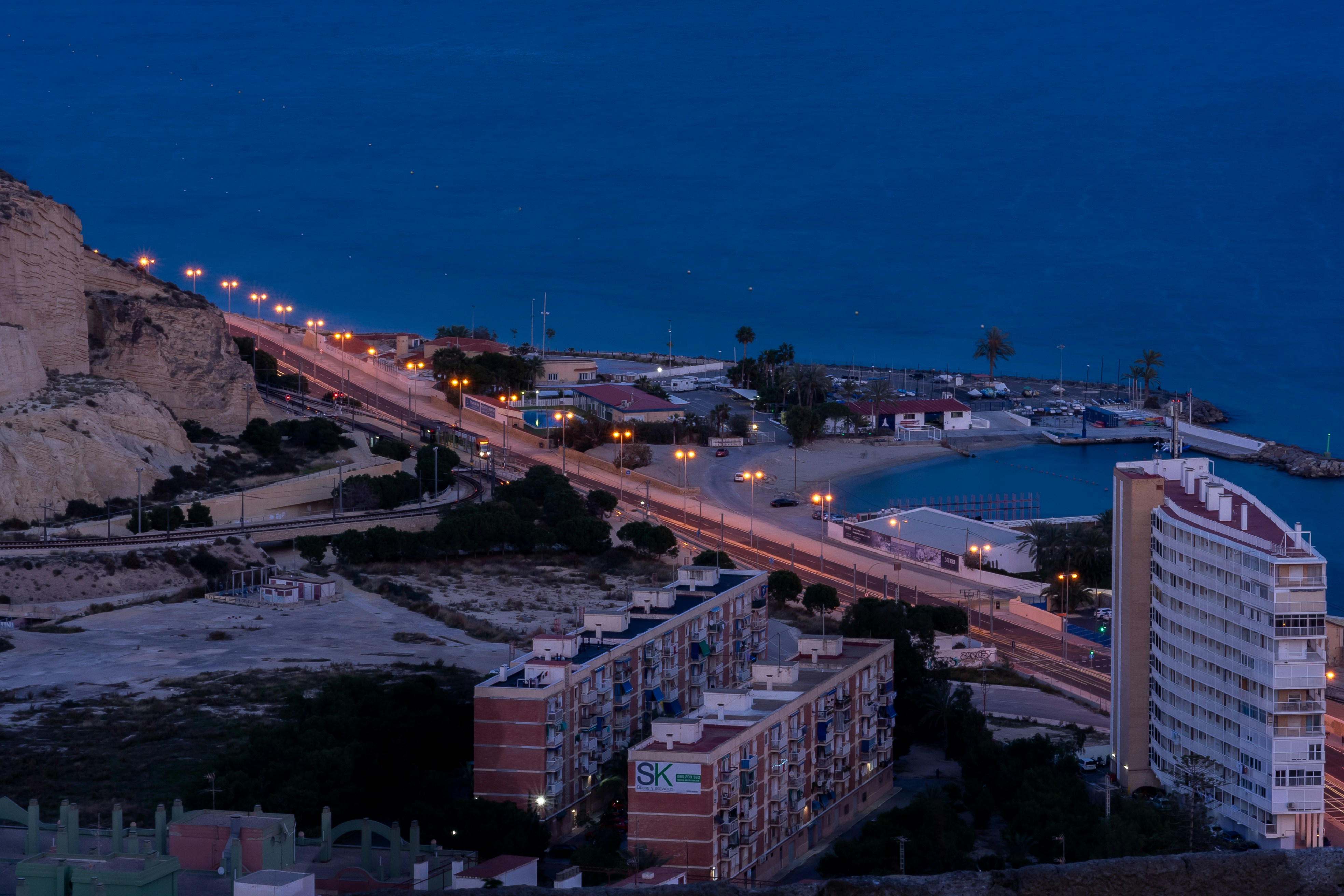 Illuminated coastal road winding along the shoreline at dusk, with nearby buildings and a tranquil harbor. 