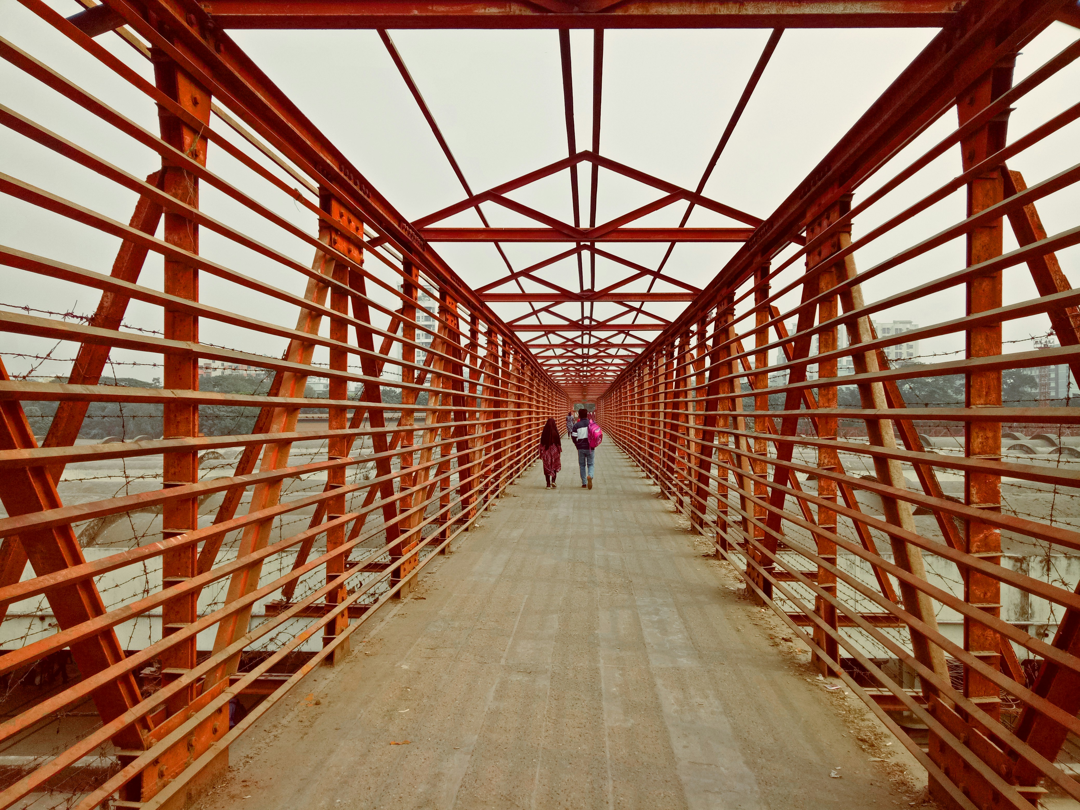 Two figures walking along a modern pedestrian bridge with striking red latticework against a muted background.