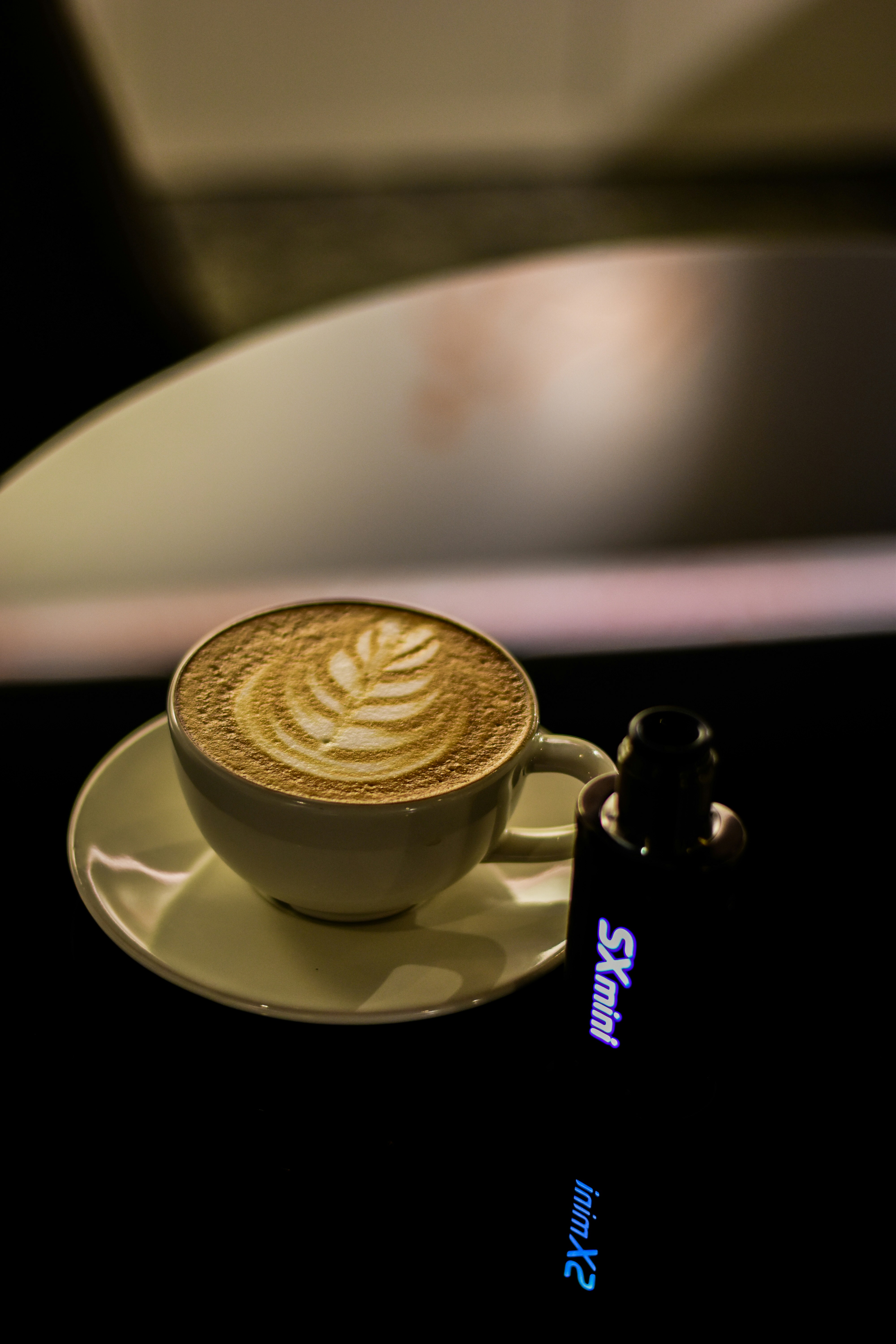 Latte art featuring a delicate leaf design in a white cup on a saucer, accompanied by a sleek vape device. The setting exudes a cozy café atmosphere.