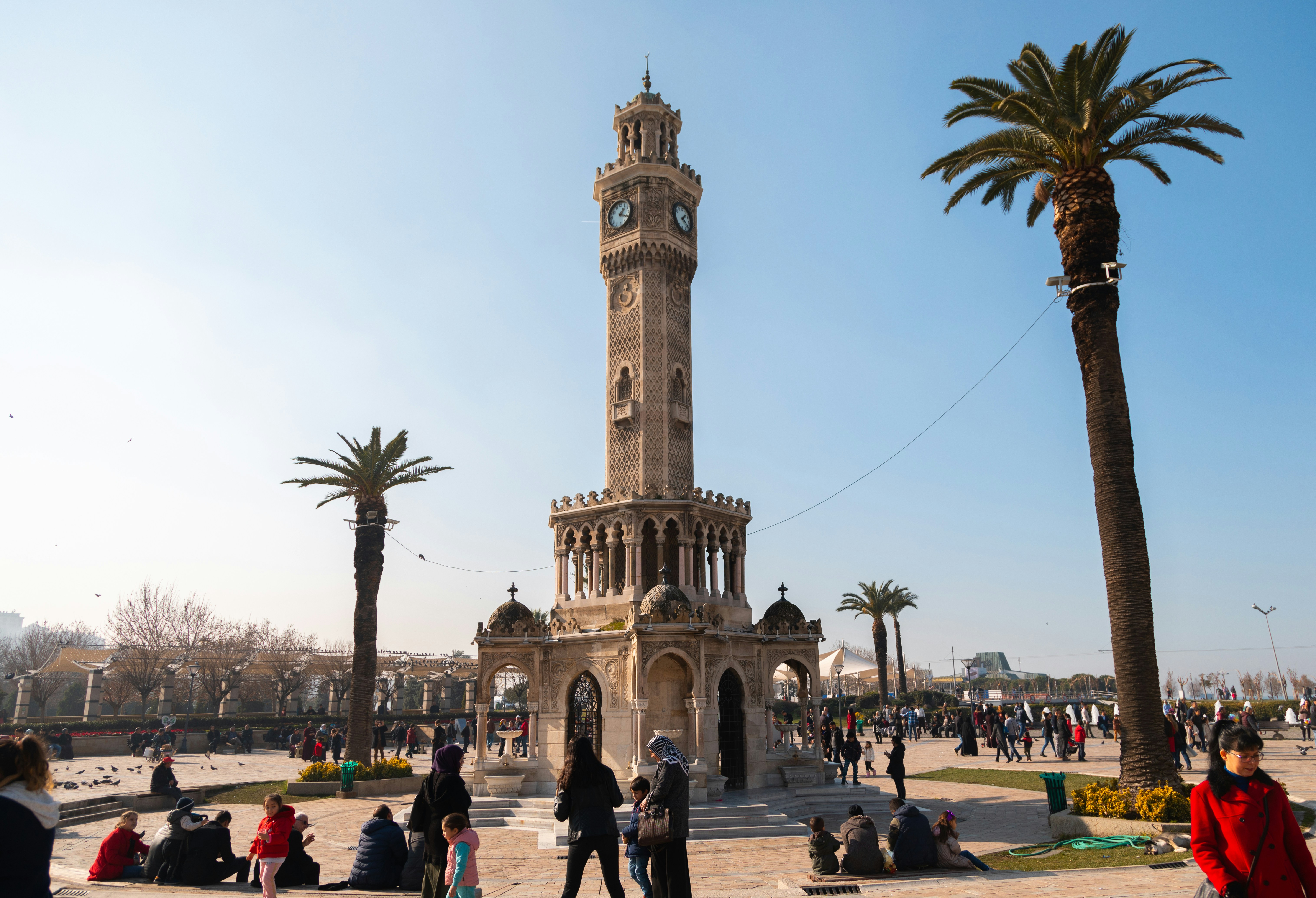 Historic clock tower surrounded by people in a vibrant city square under a clear blue sky.