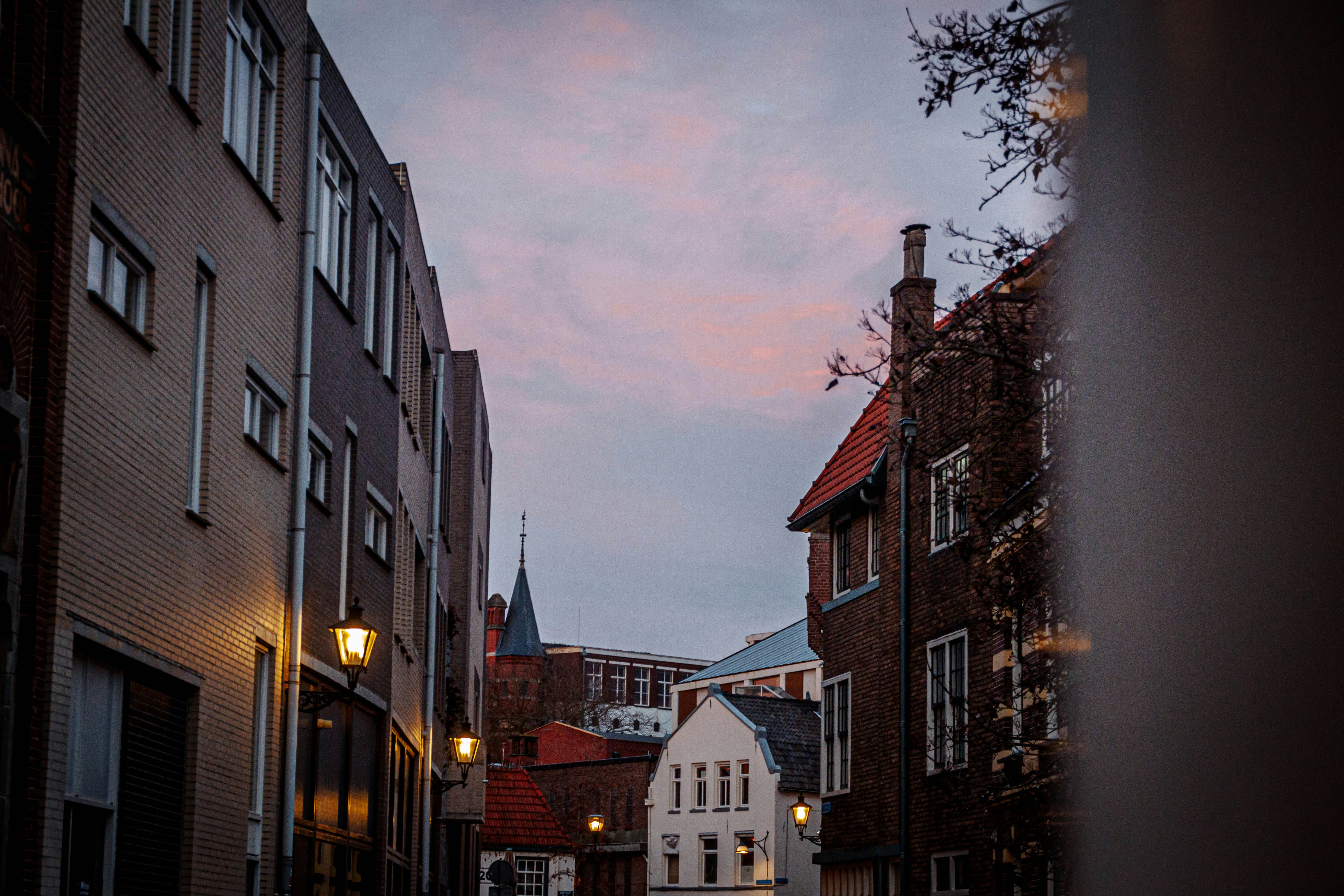 Narrow European street with warm-hued brick buildings and glowing street lamps at dusk.