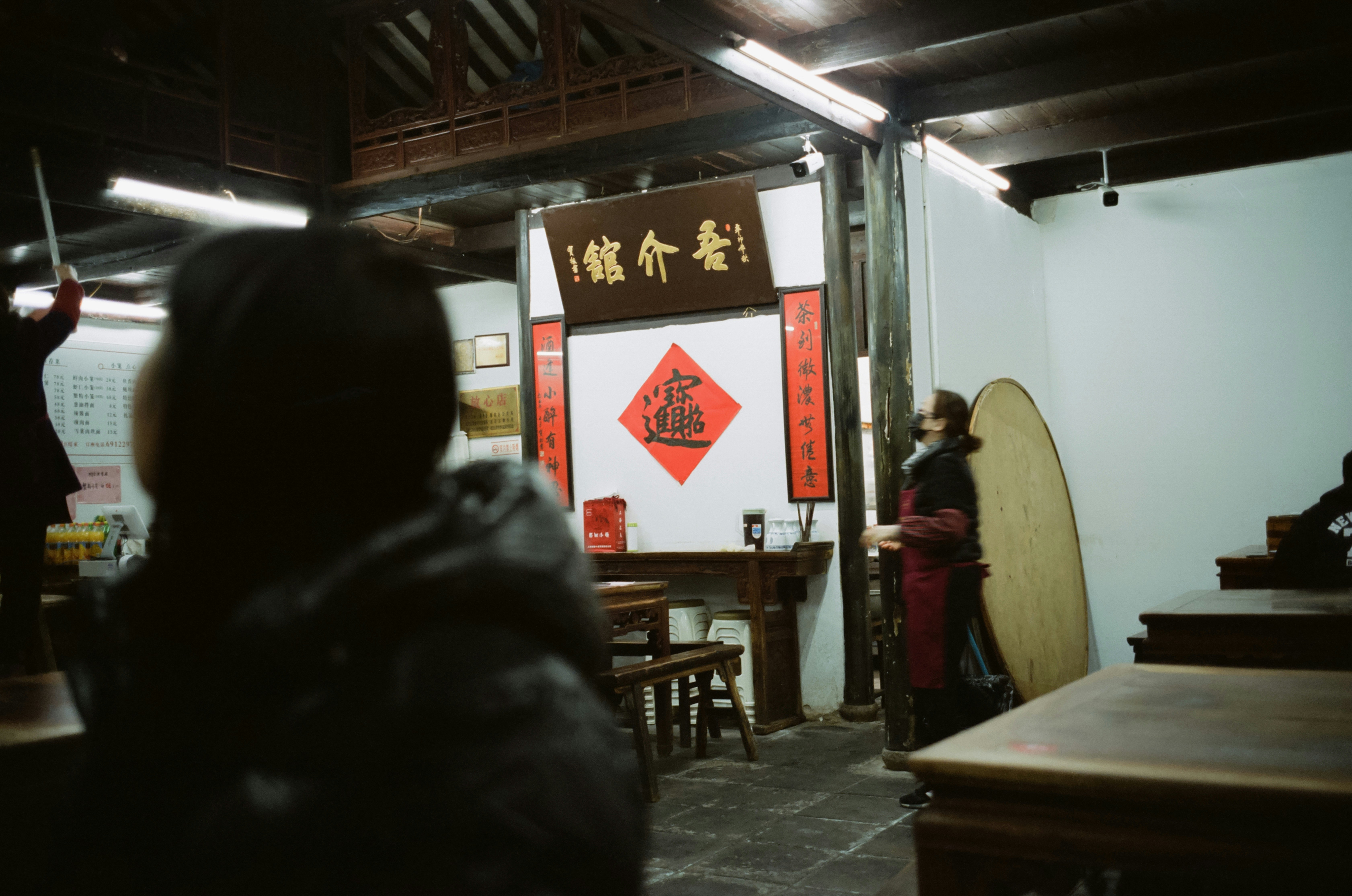 Interior of a teahouse featuring traditional decor, with patrons engaged in conversation. A striking red calligraphy piece adorns the wall.