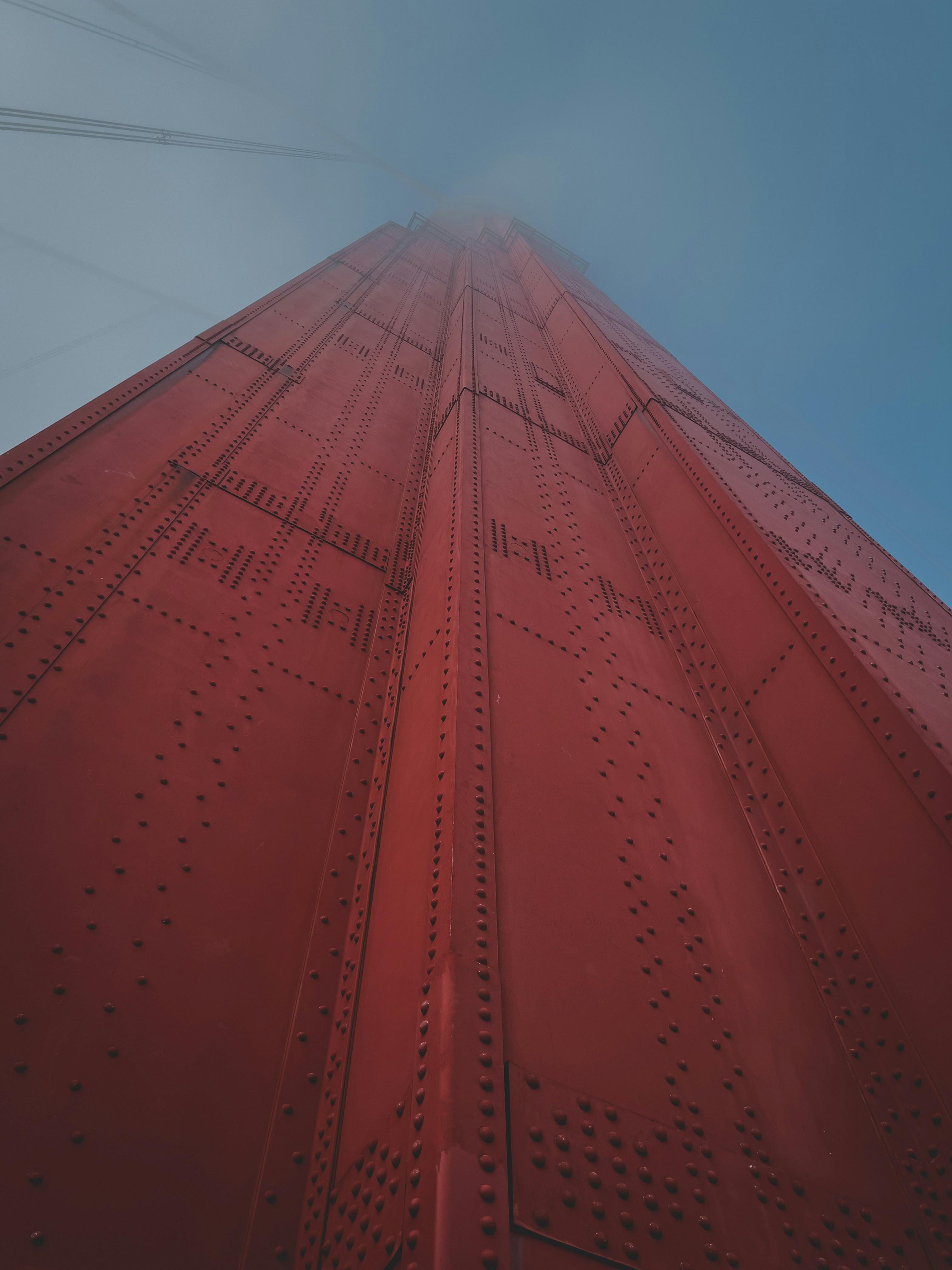 Apex of Red Rivets: Upward Gaze at a Fog-Washed Bridge Tower