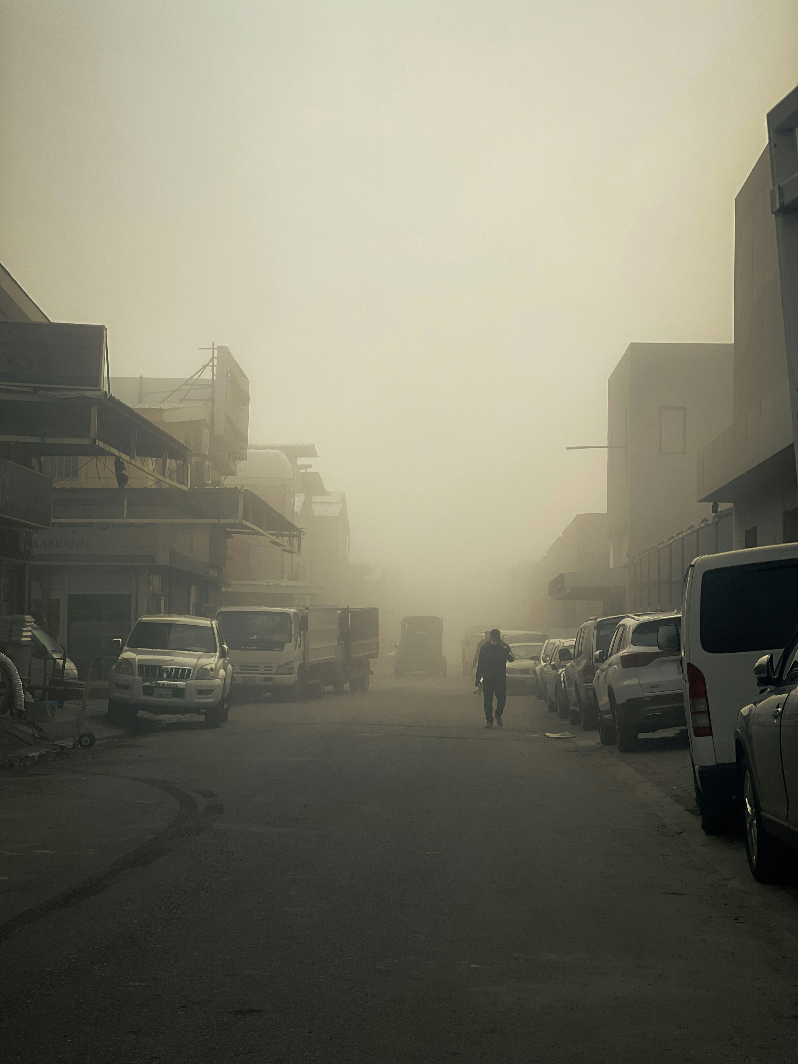 A lone figure walks down a deserted street shrouded in thick fog, with vehicles partially obscured in the mist. The atmosphere evokes a sense of mystery and solitude.