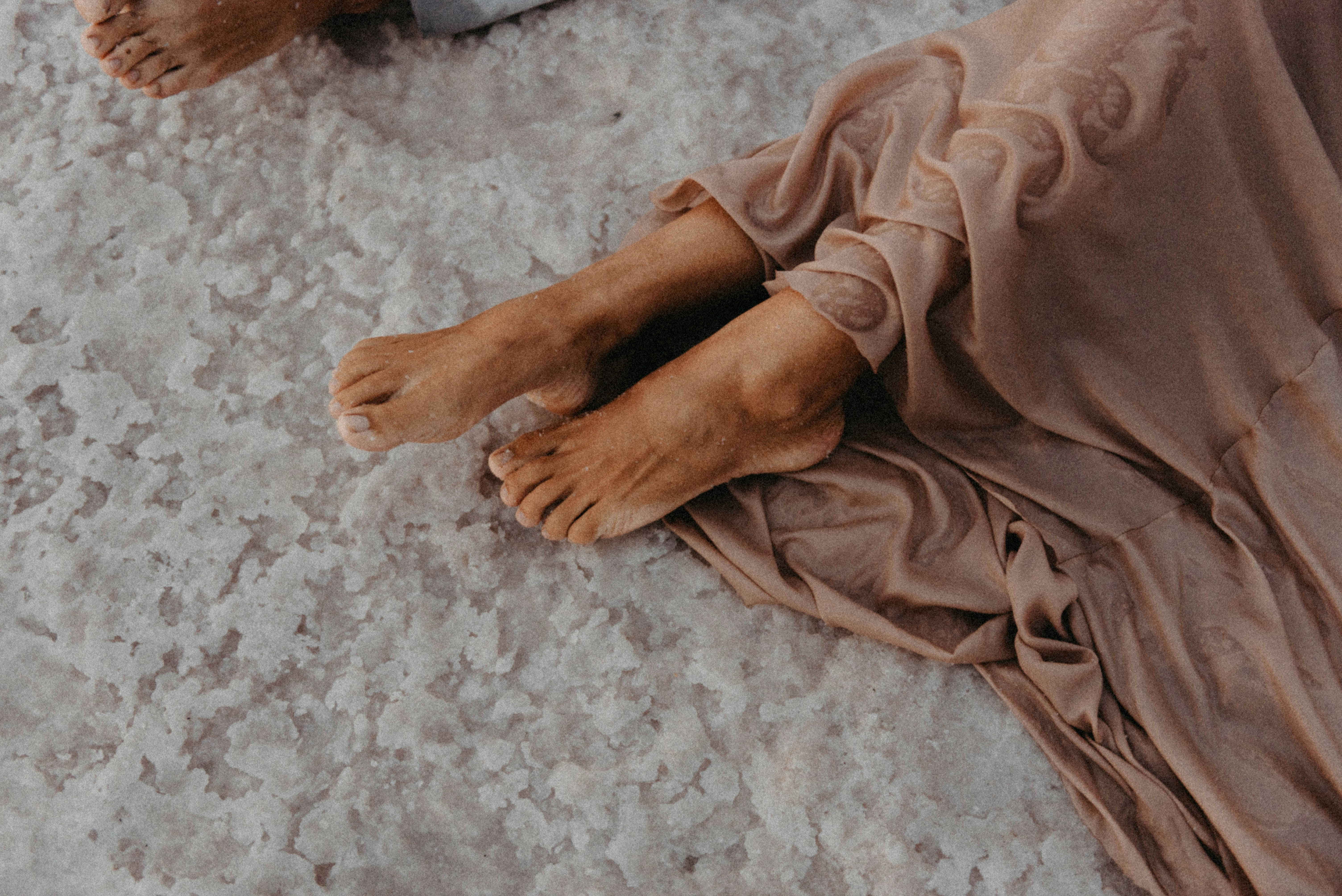 Bare feet resting on a textured surface, draped in soft fabric that flows gracefully. The interplay of skin tones and materials creates a serene atmosphere.