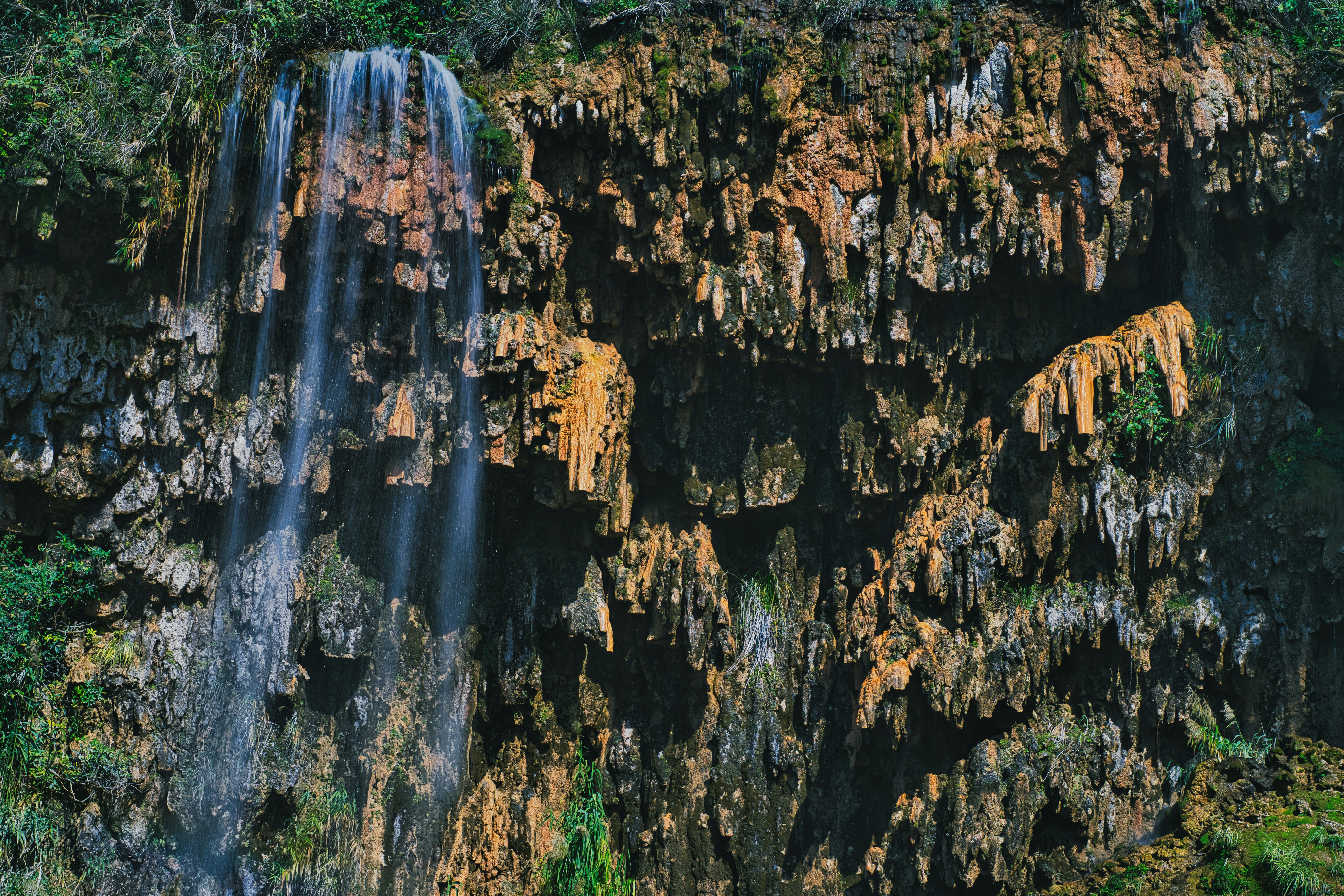 A waterfall cascading down a rugged, mineral-rich cliff, surrounded by vibrant greenery and unique rock formations.