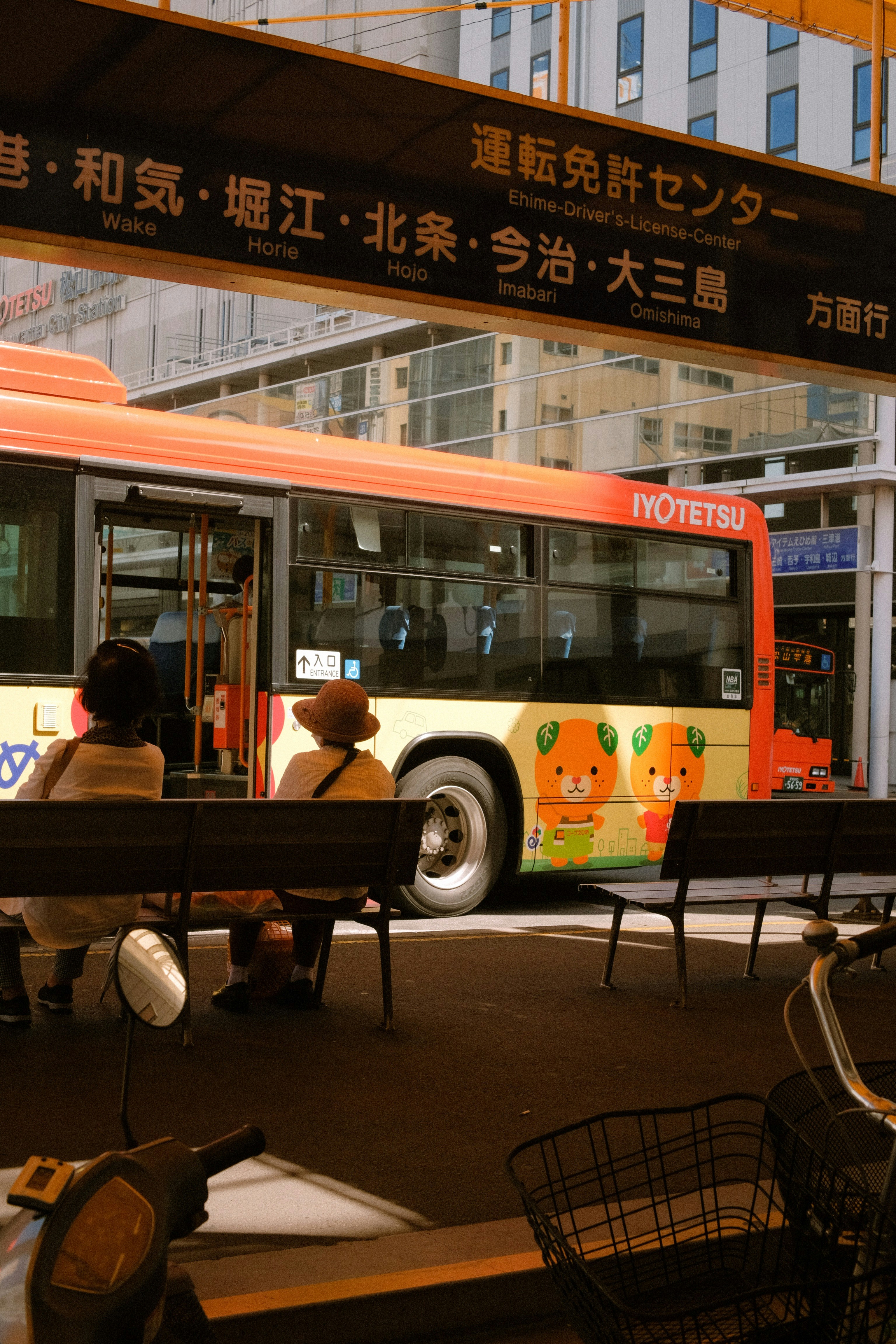 Colorful city bus parked at a transit center with passengers waiting on benches. The scene captures a moment of daily life in a Japanese urban setting.