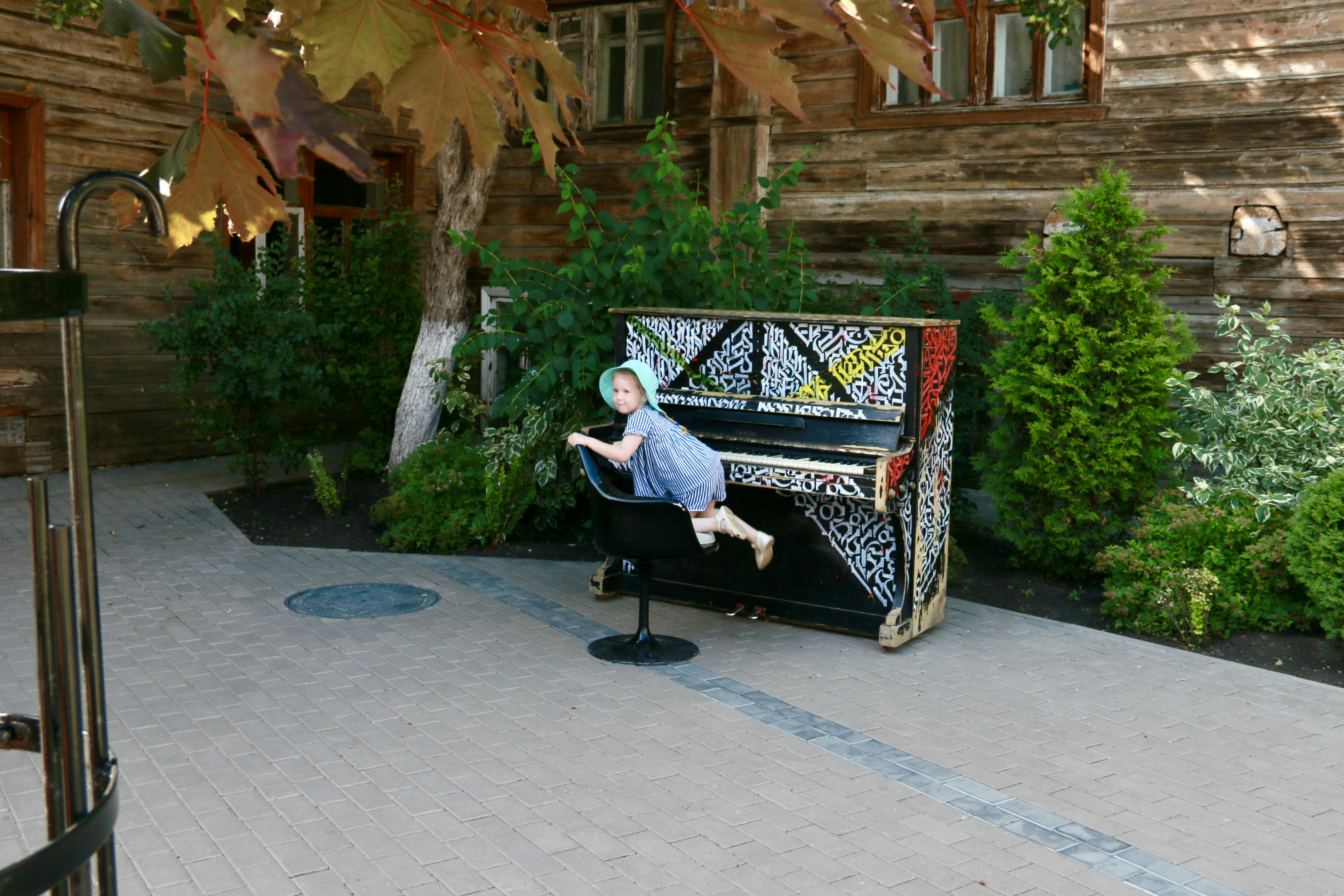 Child playfully interacting with a vibrantly painted outdoor piano against a rustic wooden house backdrop.