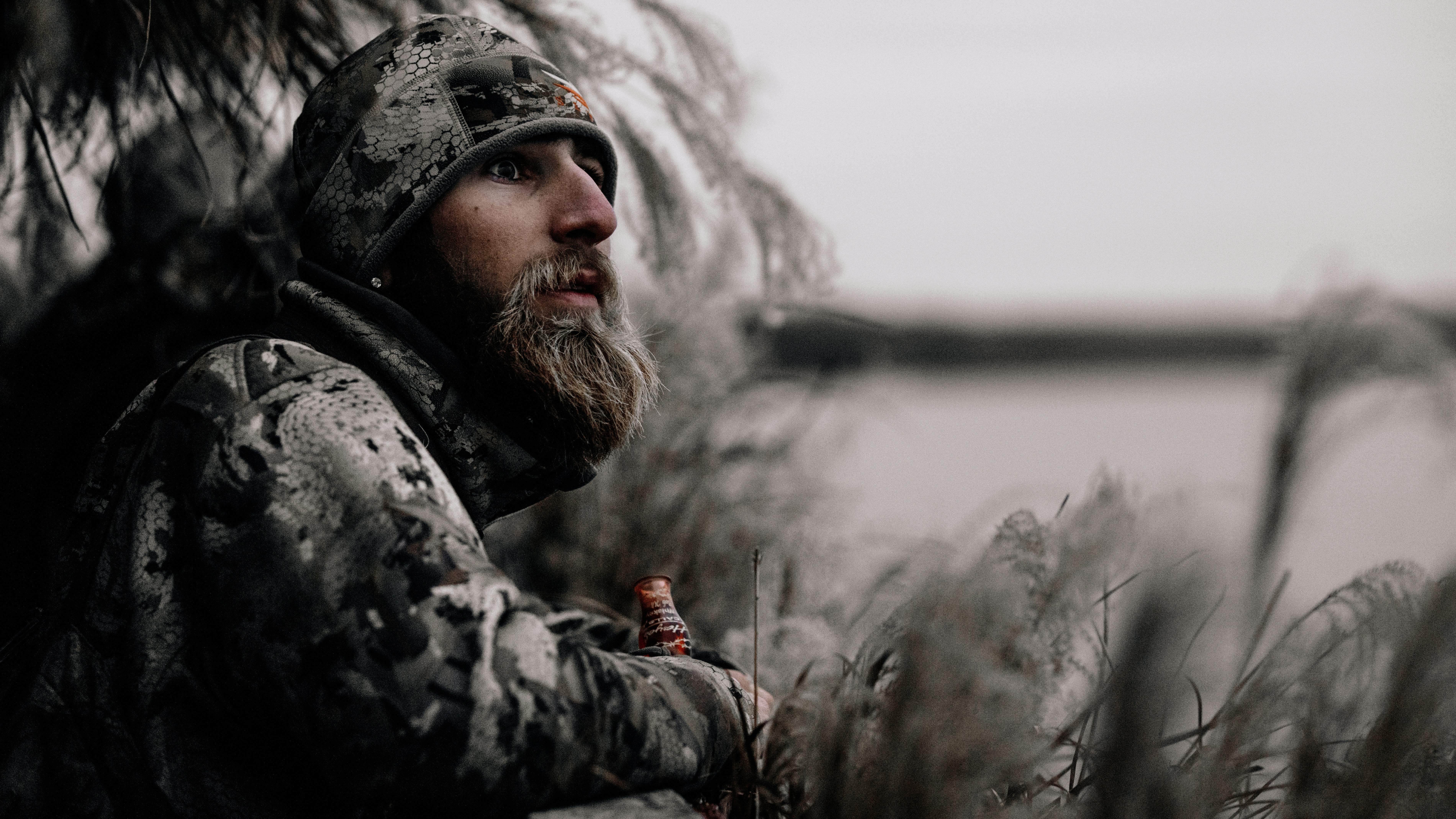 Camouflaged figure with a beard sits amidst tall grasses by a lake under a gray sky.