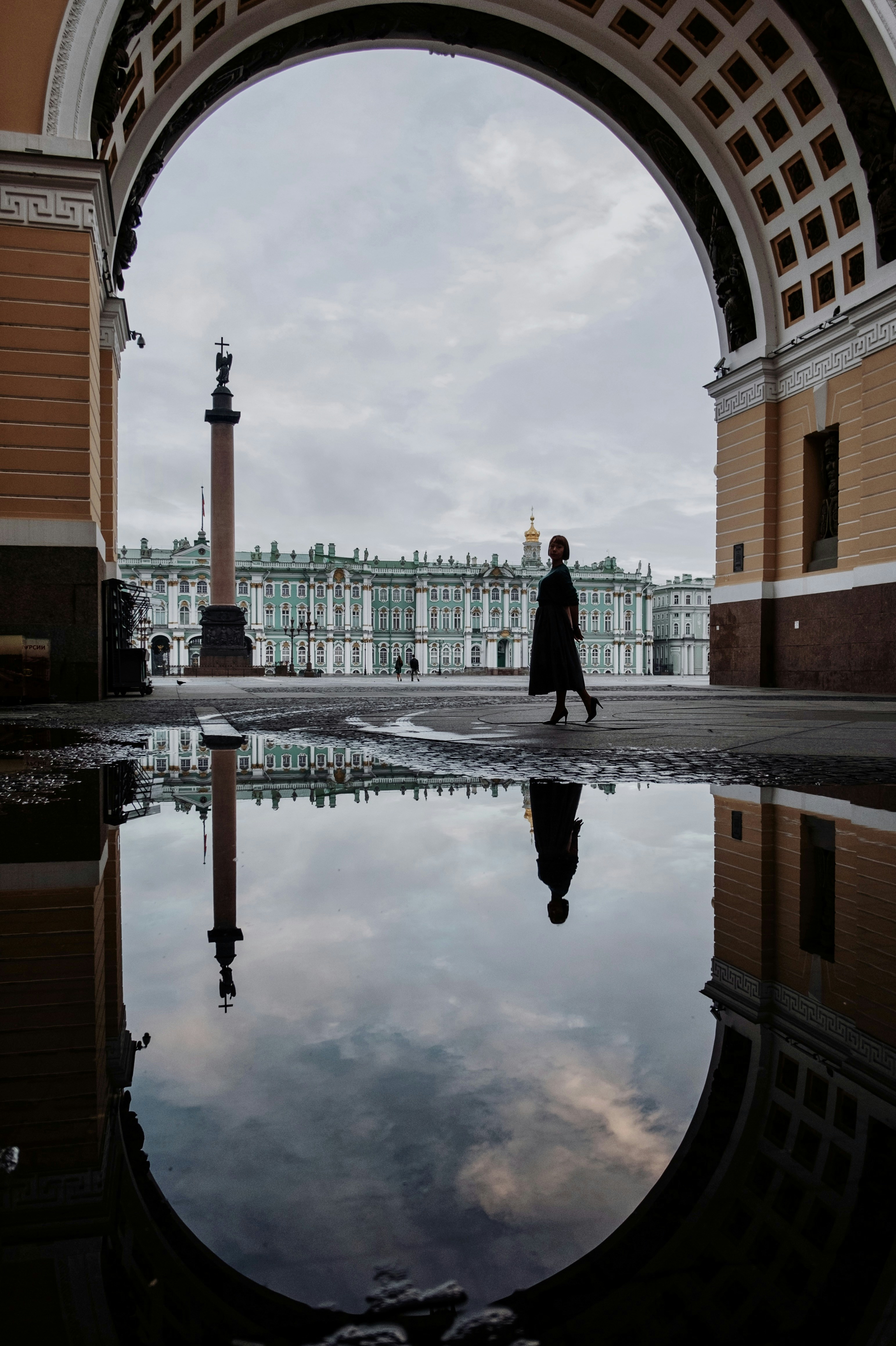 Silhouette of a person walking under an archway, with a reflective puddle capturing the grandeur of the surrounding architecture and sky.