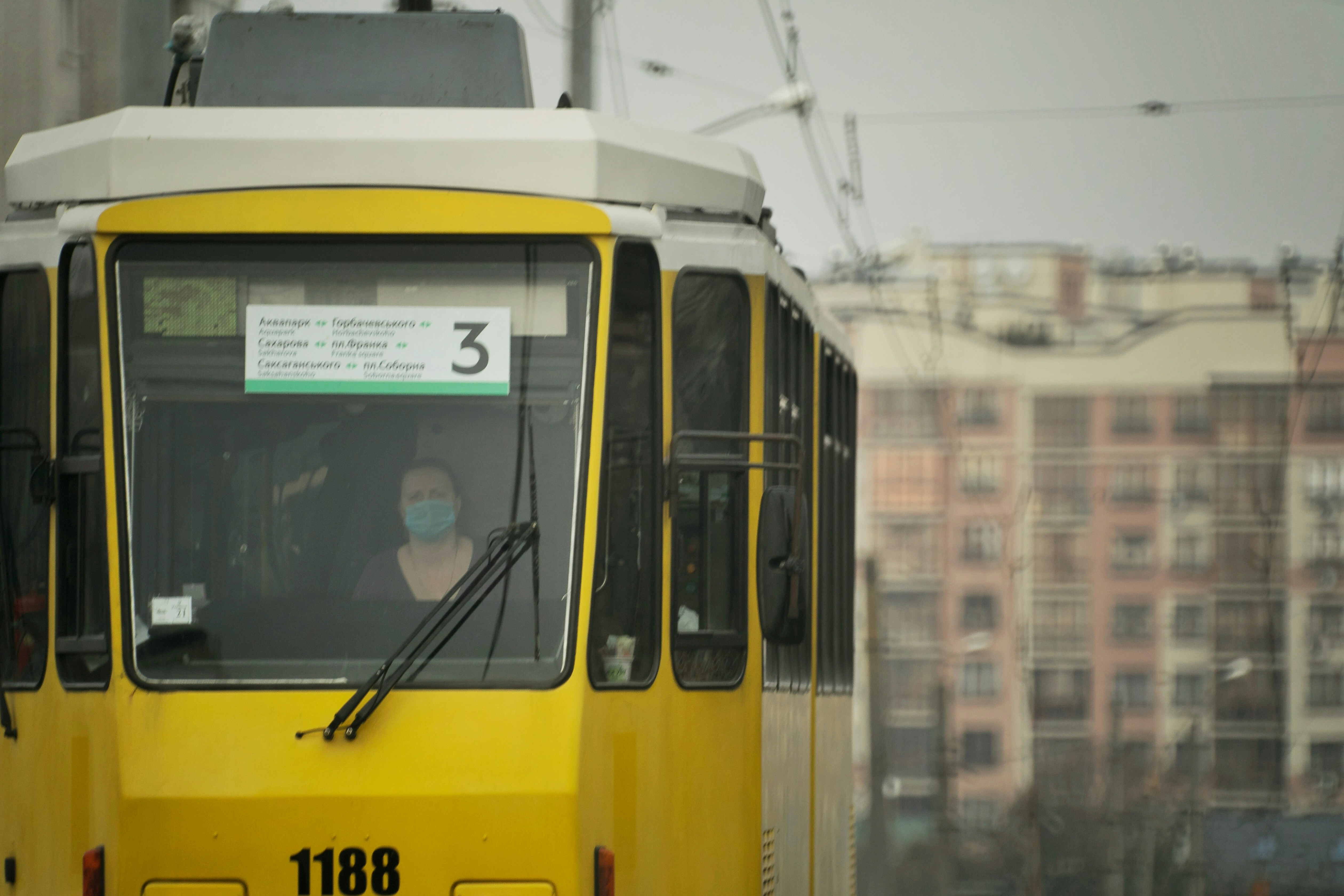 Yellow tram with a masked passenger visible through the front window, navigating a cityscape. The background features urban architecture, hinting at a bustling environment.