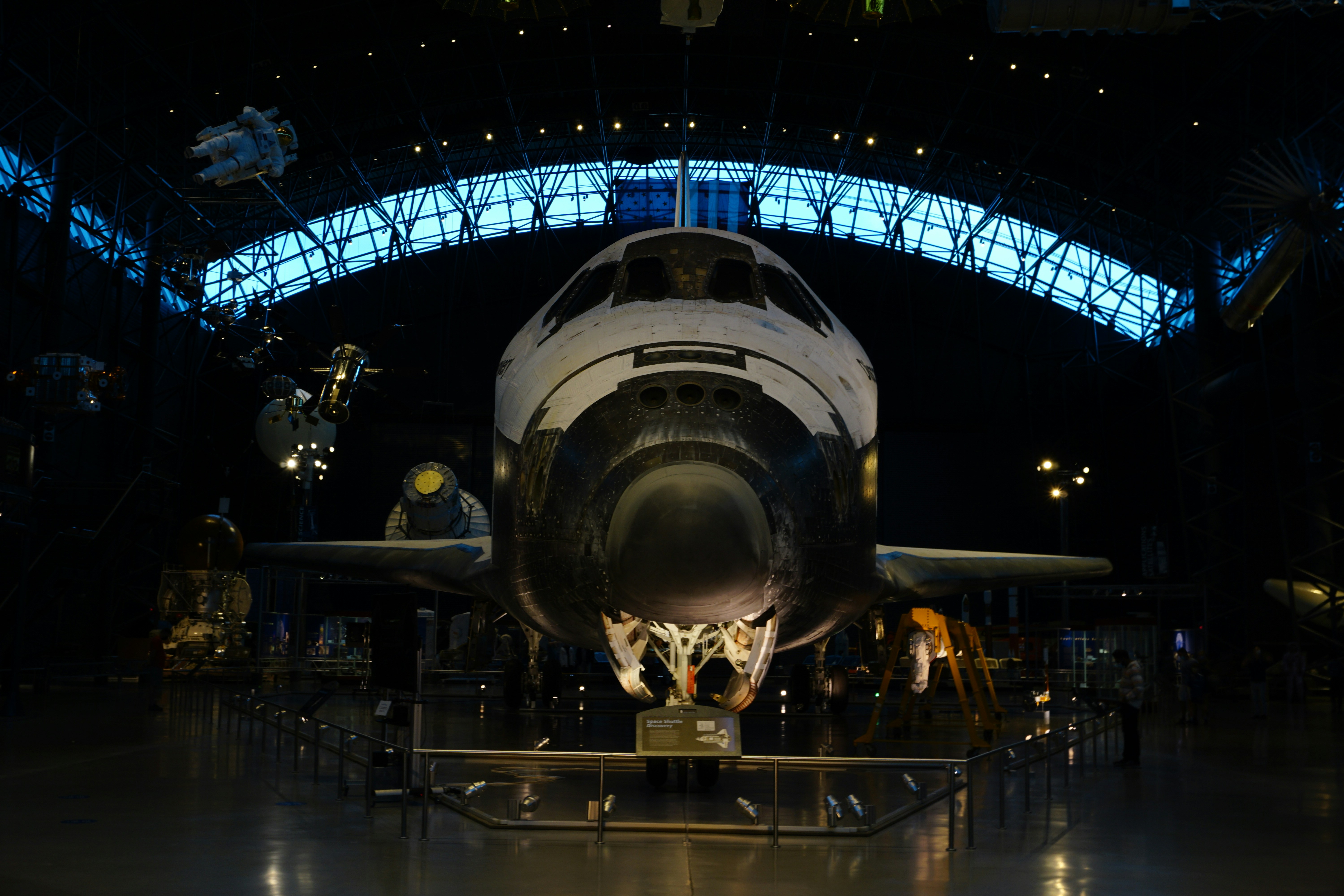 Space shuttle displayed in a dimly lit museum setting, surrounded by other aerospace artifacts.
