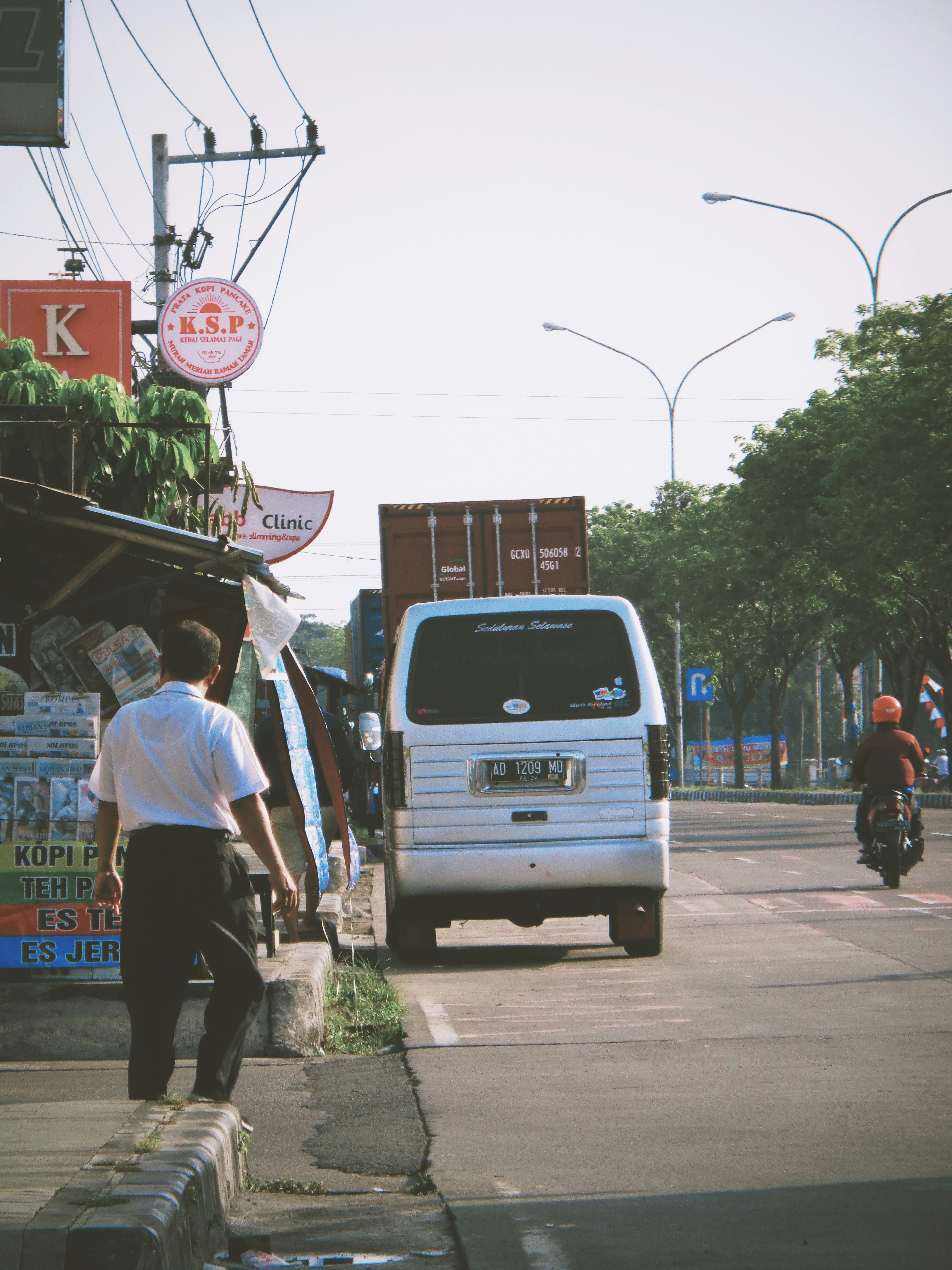 A man in a white shirt walks past a roadside stall adorned with posters, while a white van and a motorcycle navigate the street ahead. 