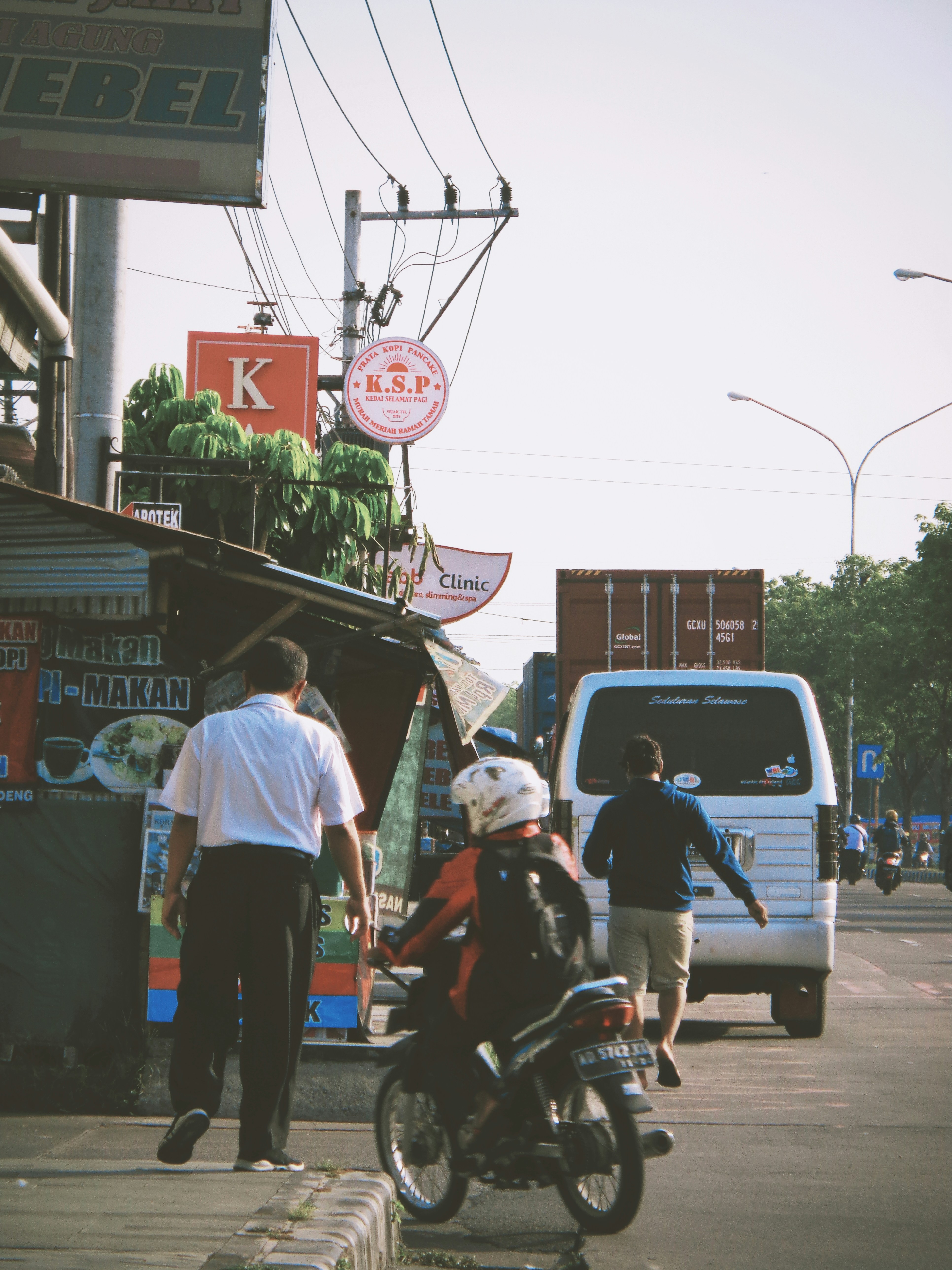 Busy street scene with a helmeted motorcyclist in the foreground, pedestrians, and a white bus among signs and storefronts along a sidewalk.