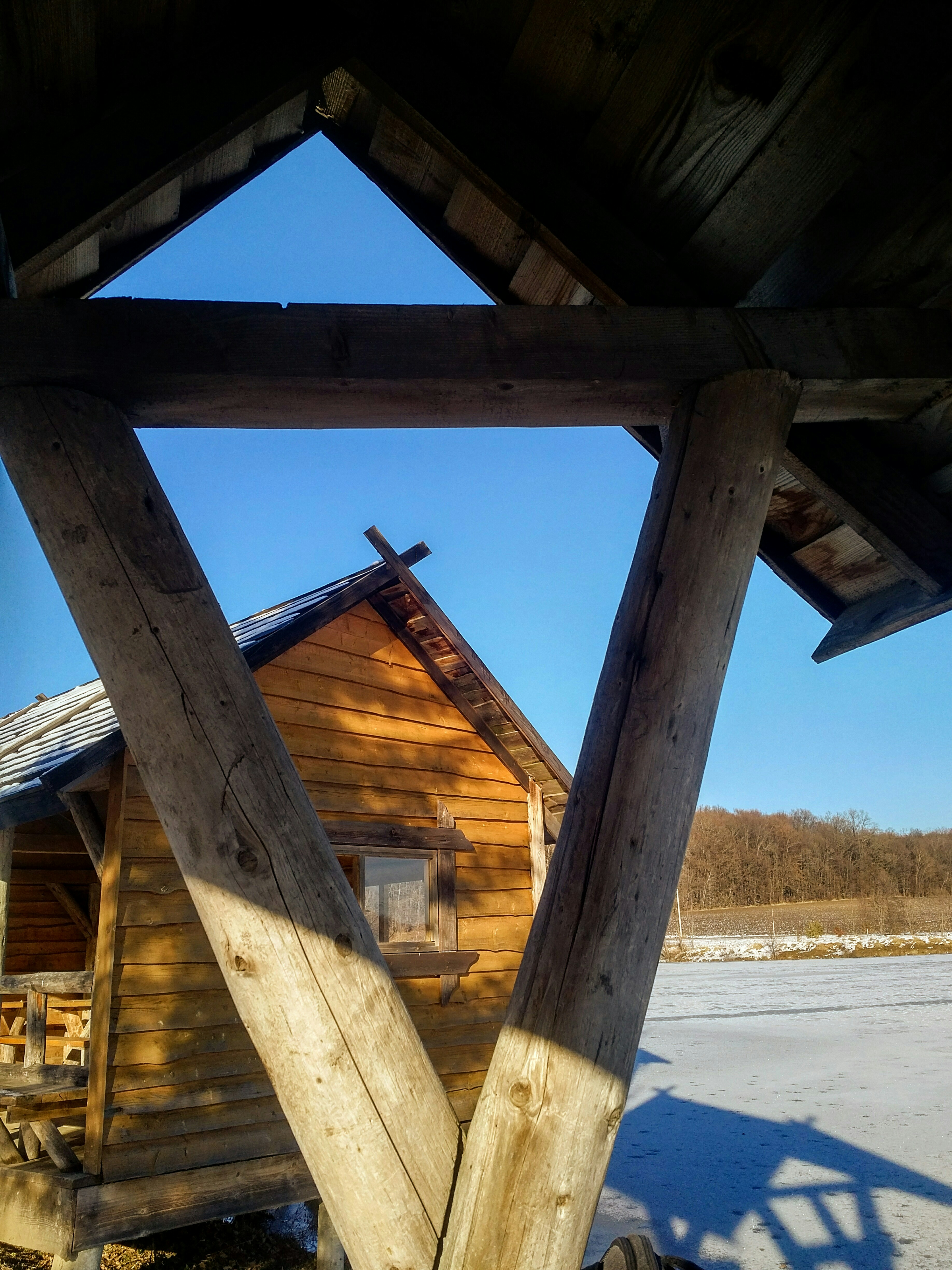 A wooden cabin framed by rustic beams under a clear blue sky, highlighting the tranquility of the countryside in winter.