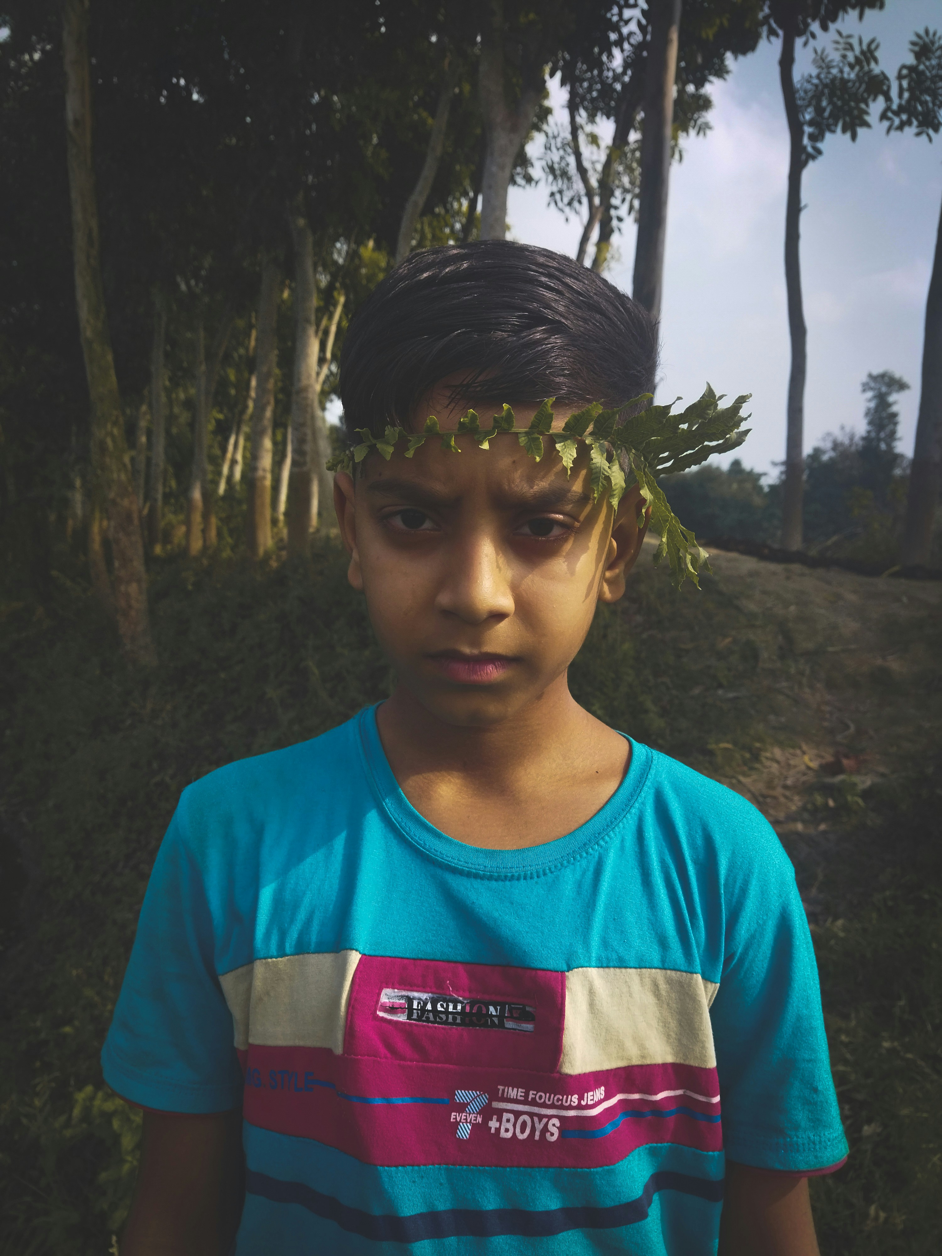 A young boy wearing a crown made of leaves stands confidently among trees, showcasing a blend of innocence and nature's beauty.