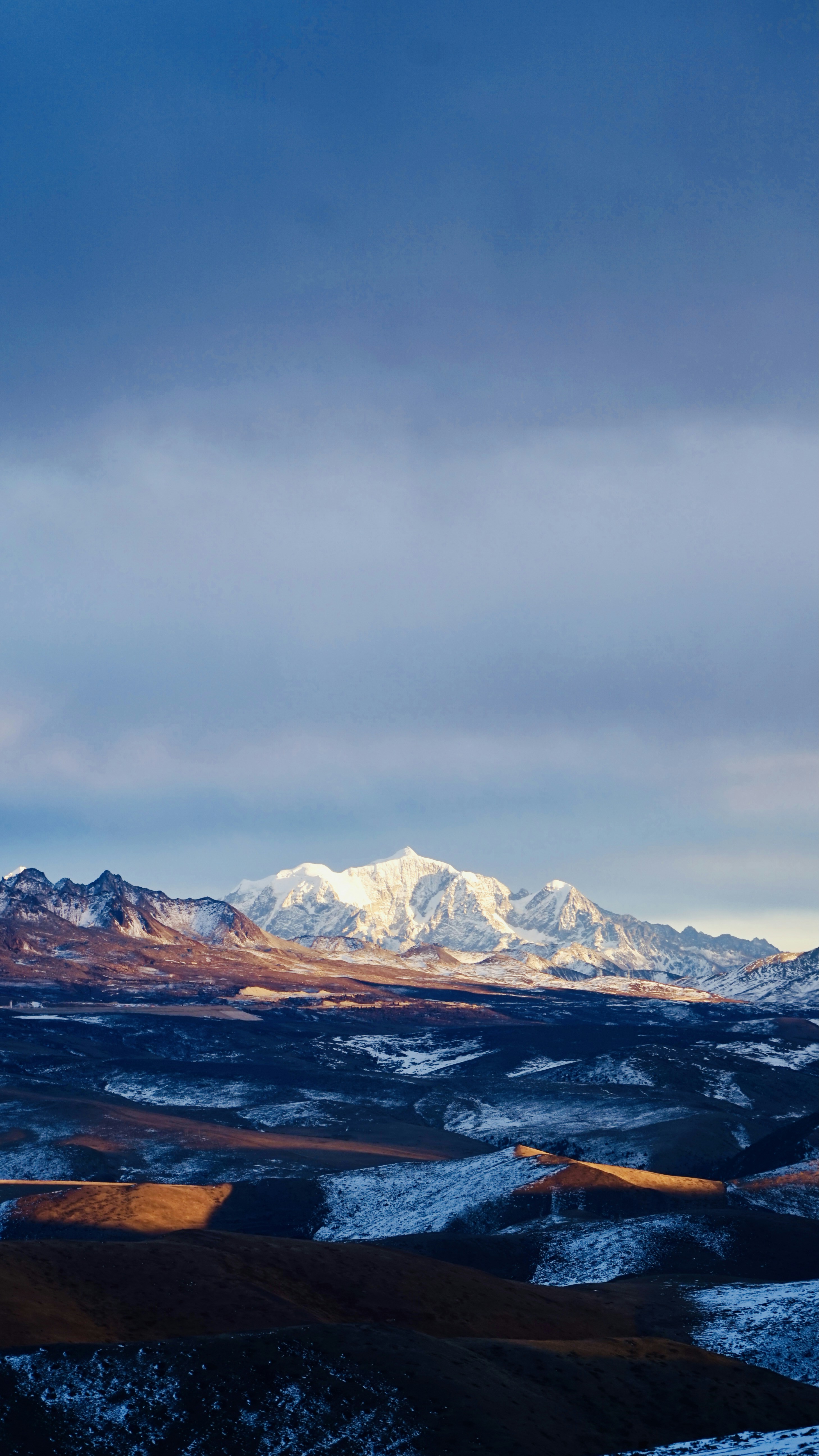 Majestic snow-capped mountains rise above a patchwork of valleys and fields, illuminated by soft winter light.