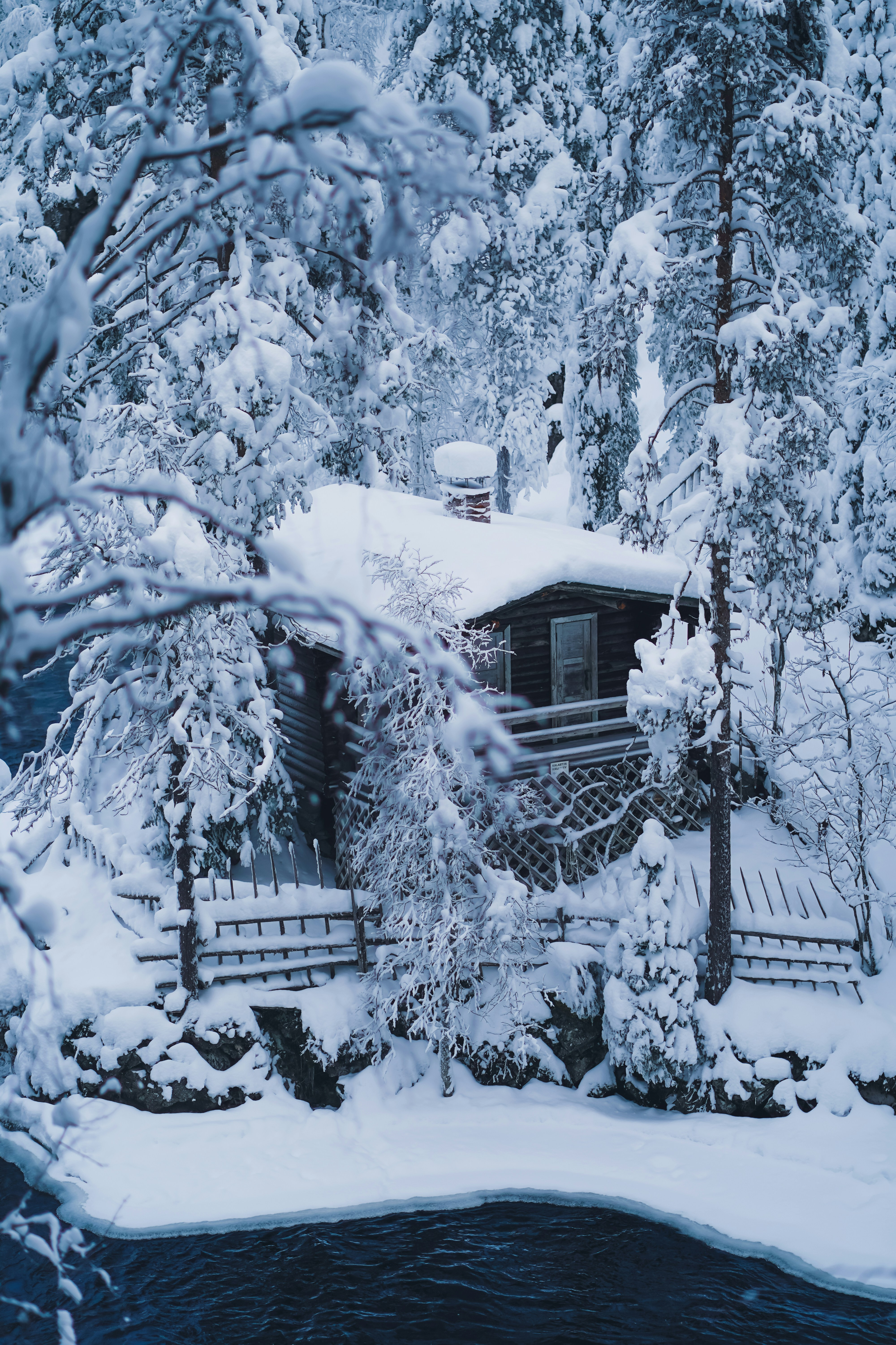 A cozy wooden cabin nestled in a snowy landscape, surrounded by frosted trees and a gently flowing river. The tranquil scene evokes a sense of peaceful solitude.