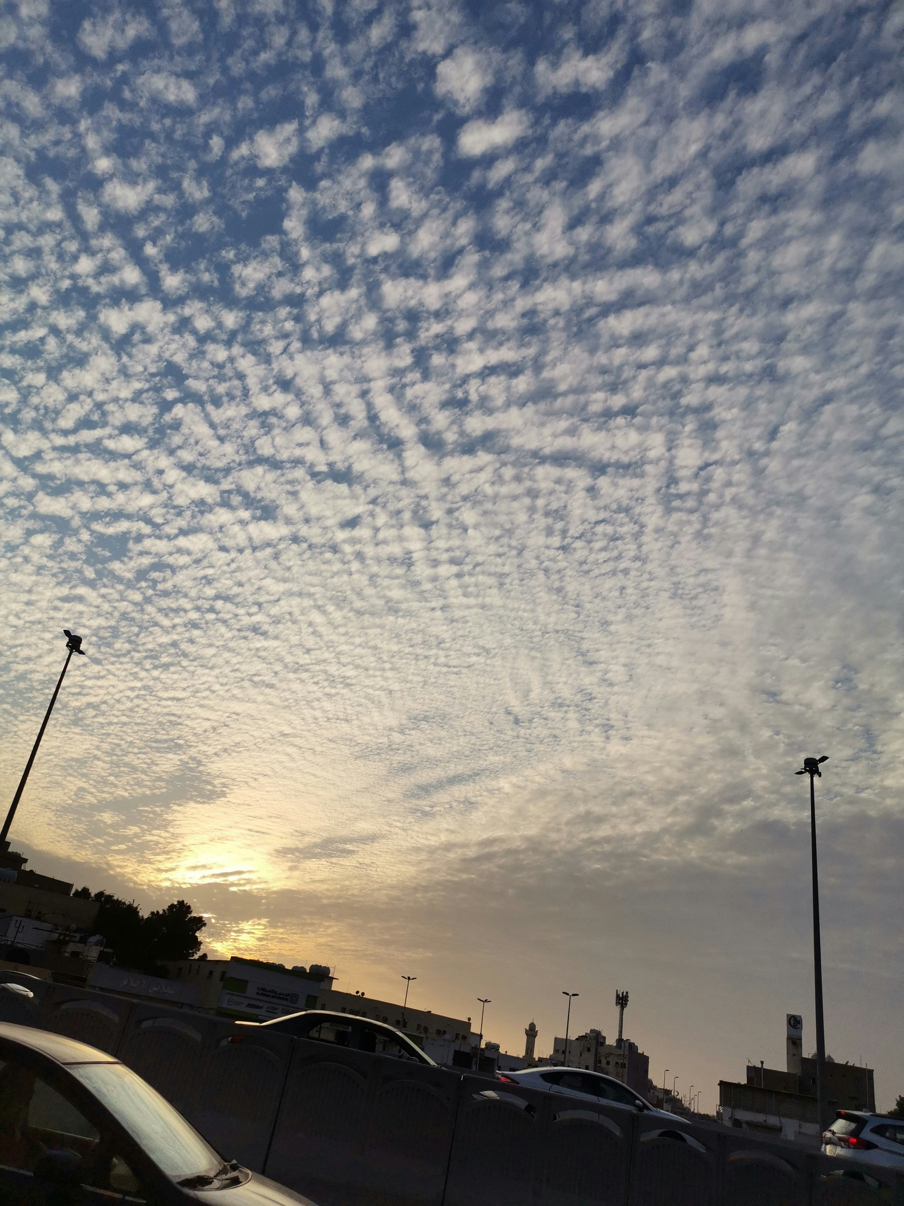 Clouds ripple across the sky as the sun dips below the horizon, casting a gentle glow. Silhouettes of streetlights and buildings frame the scene.