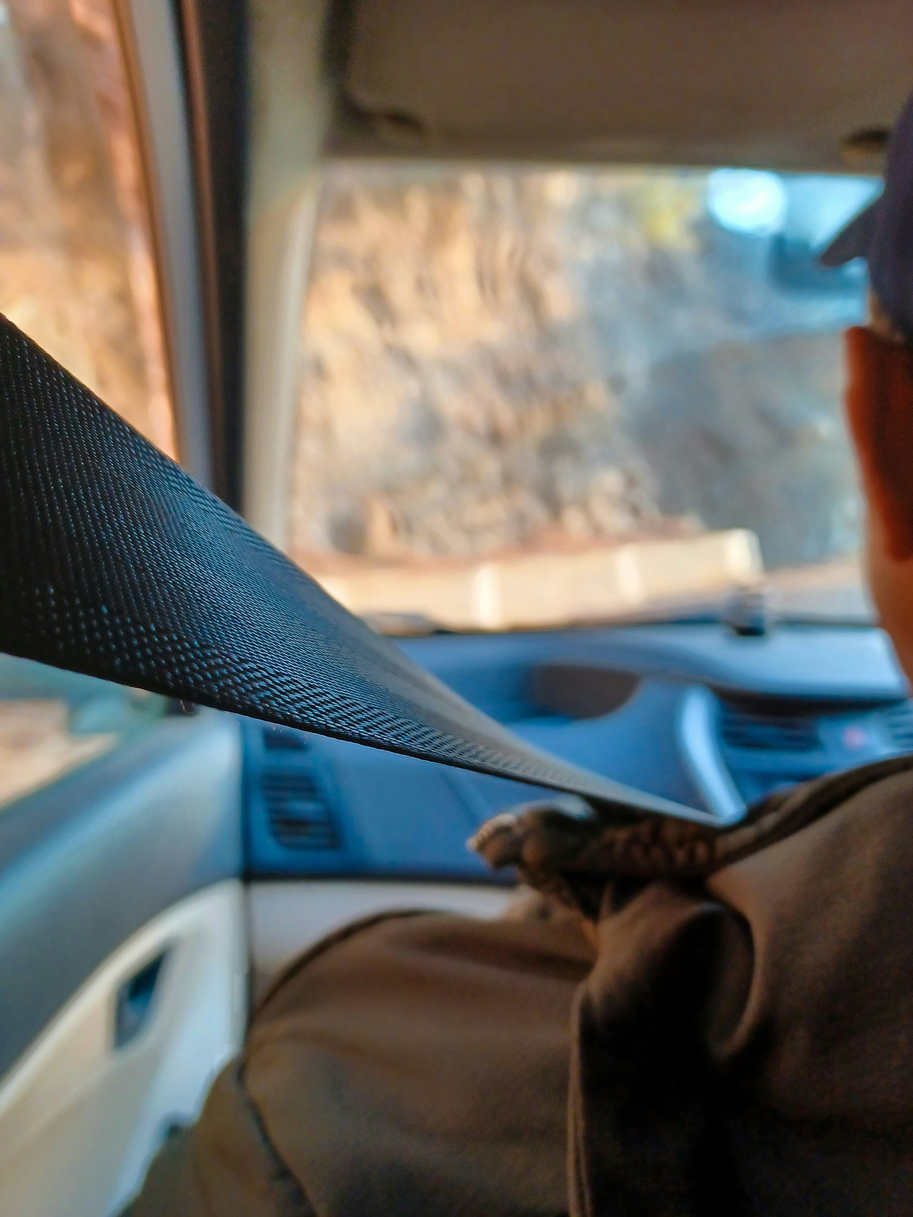 Close-up photo of a car interior featuring a diagonal seatbelt across the foreground, with the blurred road beyond implying motion.