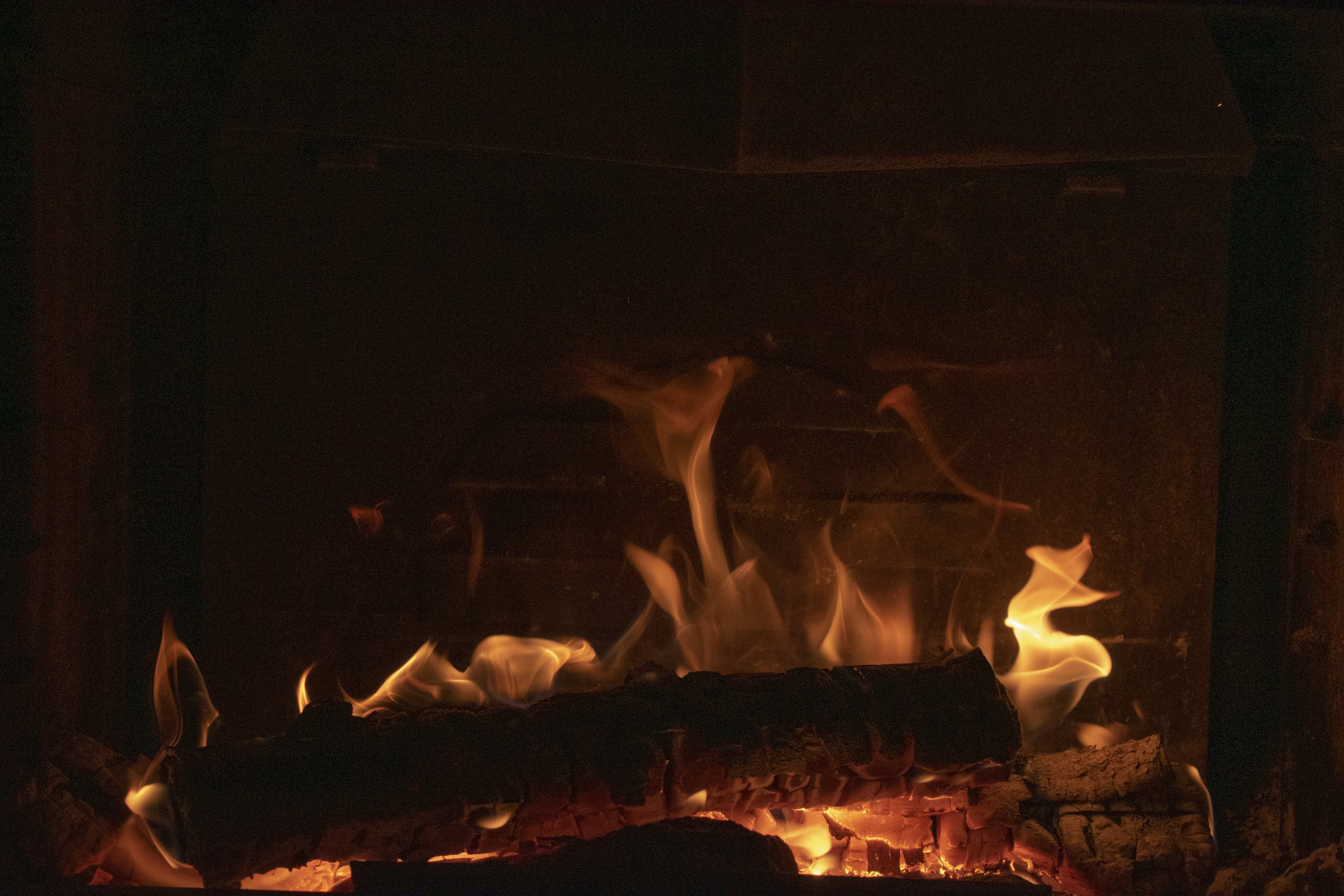 Flickering flames rise from glowing embers in a cozy fireplace, casting a warm glow in a dimly lit room.