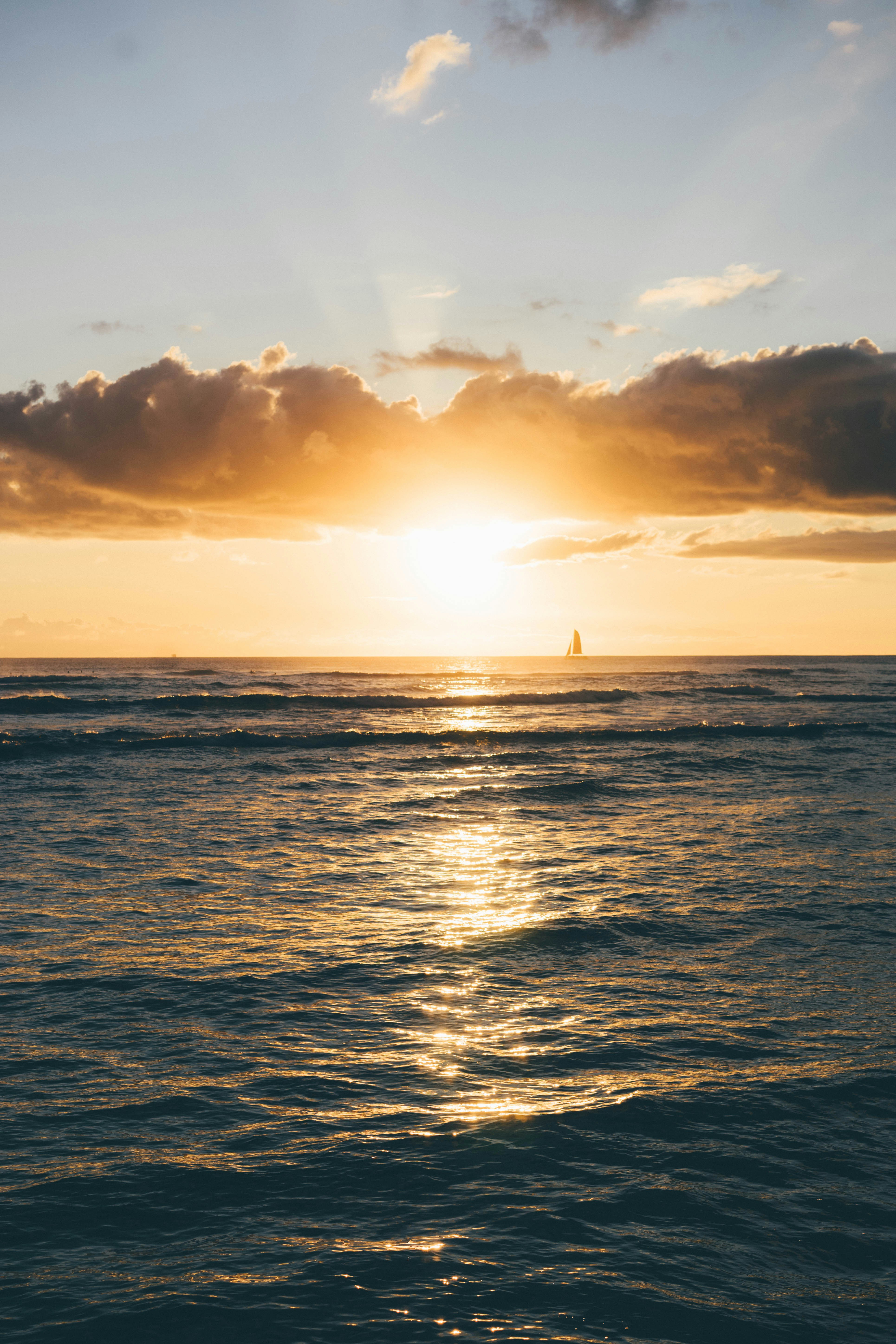 A sailboat glides across the shimmering ocean as the sun sets, casting golden reflections on the water's surface.