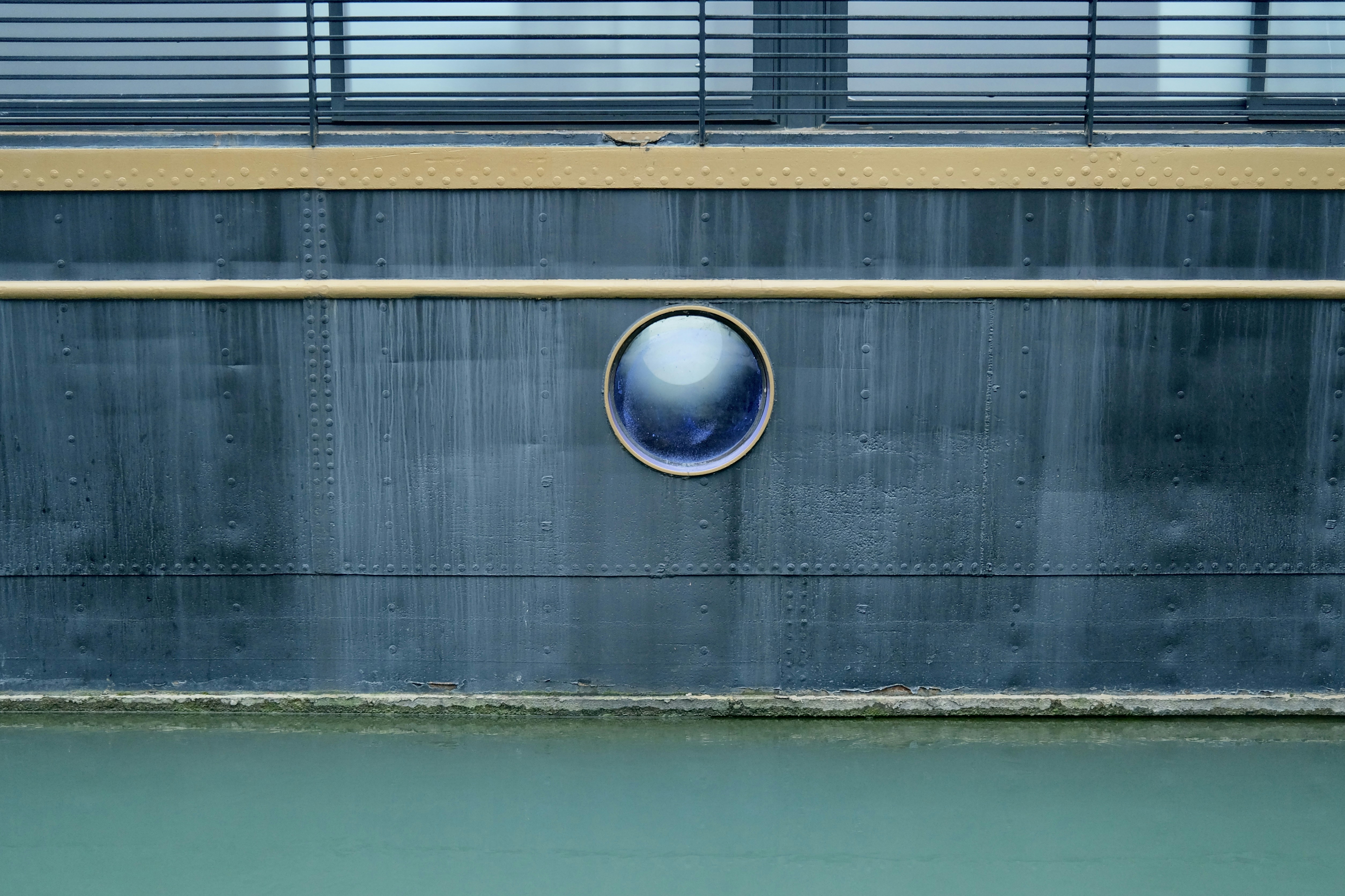 Circular porthole revealing reflections on a ship's hull, framed by a contrasting gold trim against a textured blue surface. 